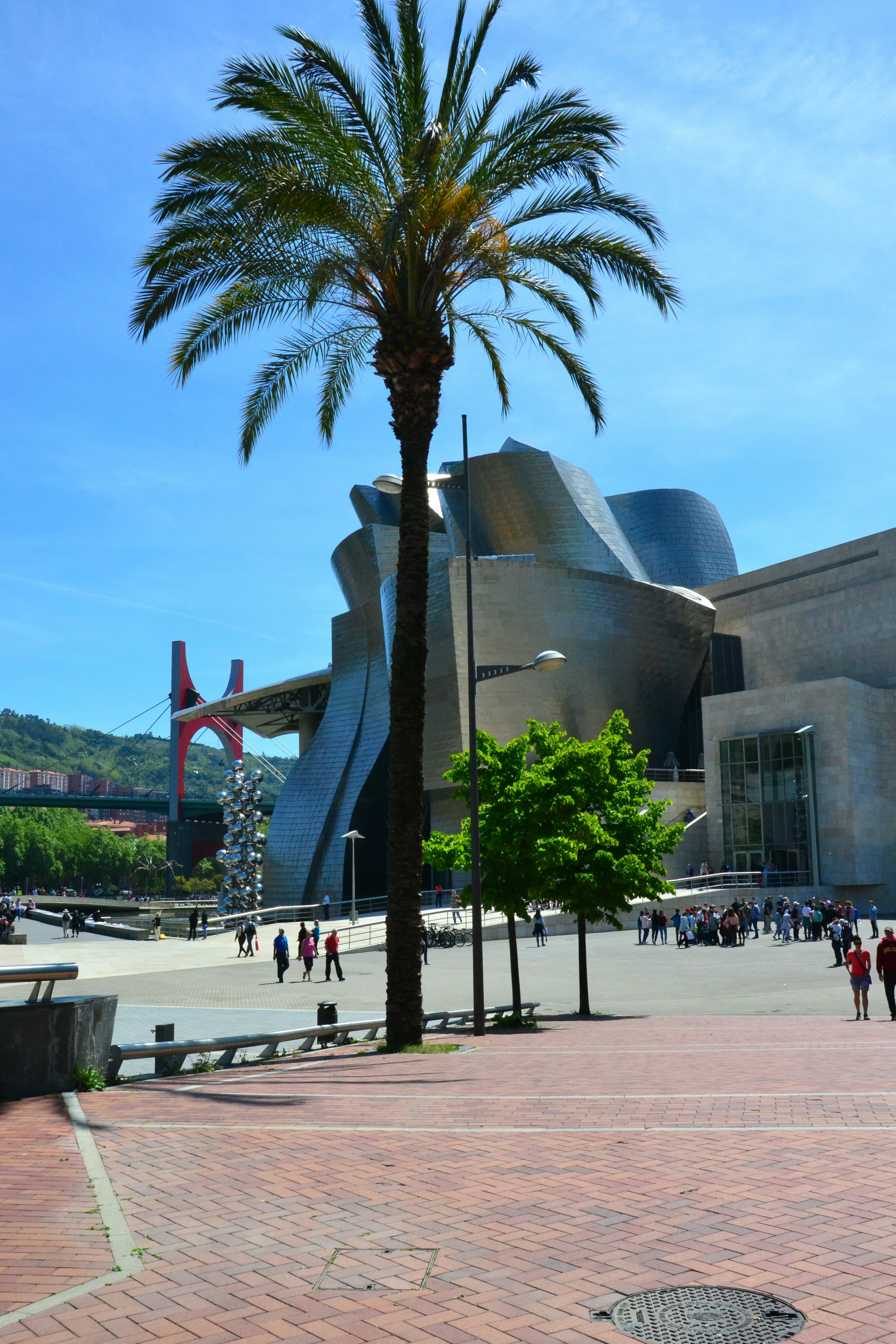 Palm tree framing the iconic Guggenheim Museum in Bilbao, showcasing a blend of organic and modern design elements. Visitors stroll through the vibrant plaza.
