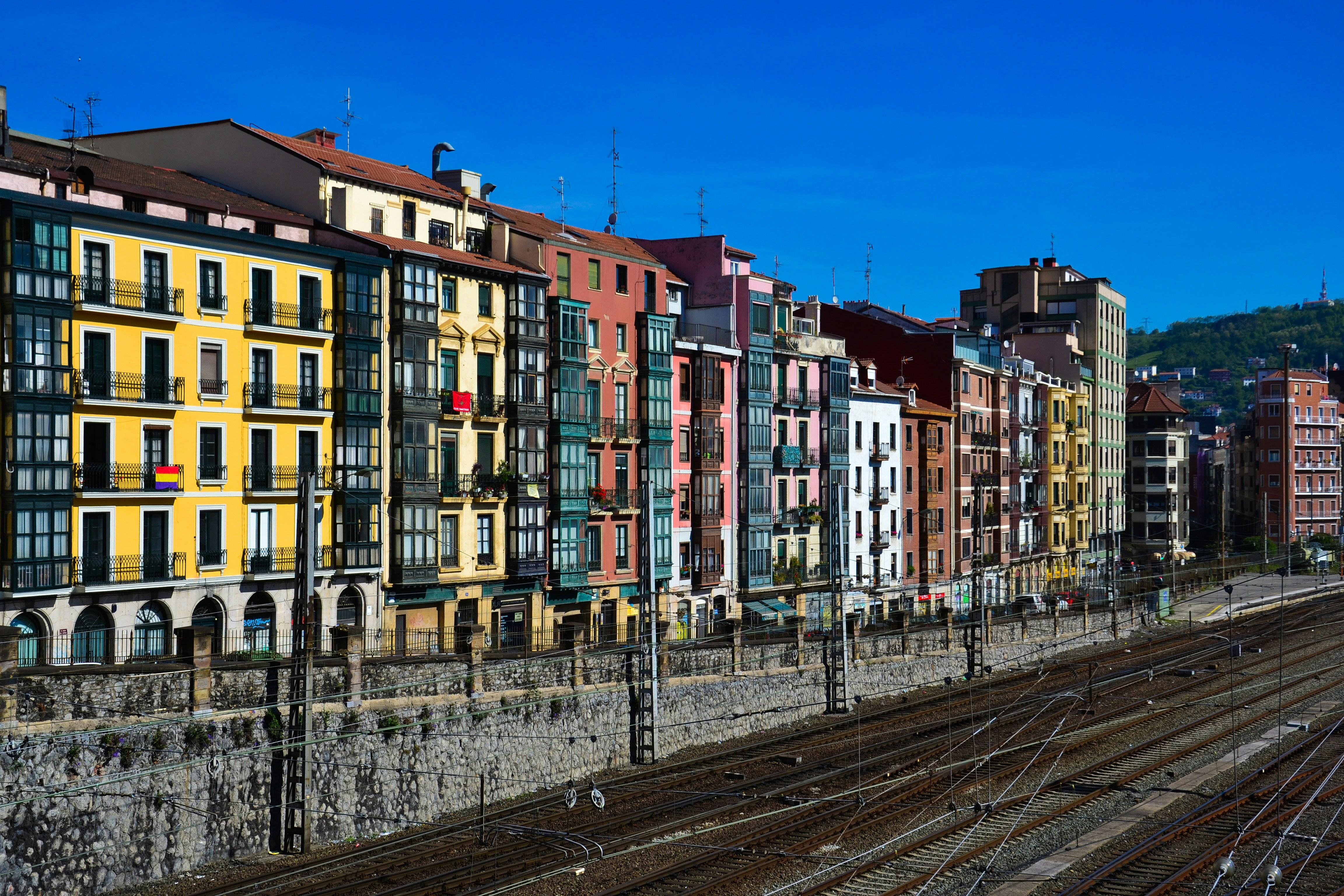 Colorful buildings line the railway tracks in a bustling urban setting, showcasing a mix of architectural styles under a clear blue sky.