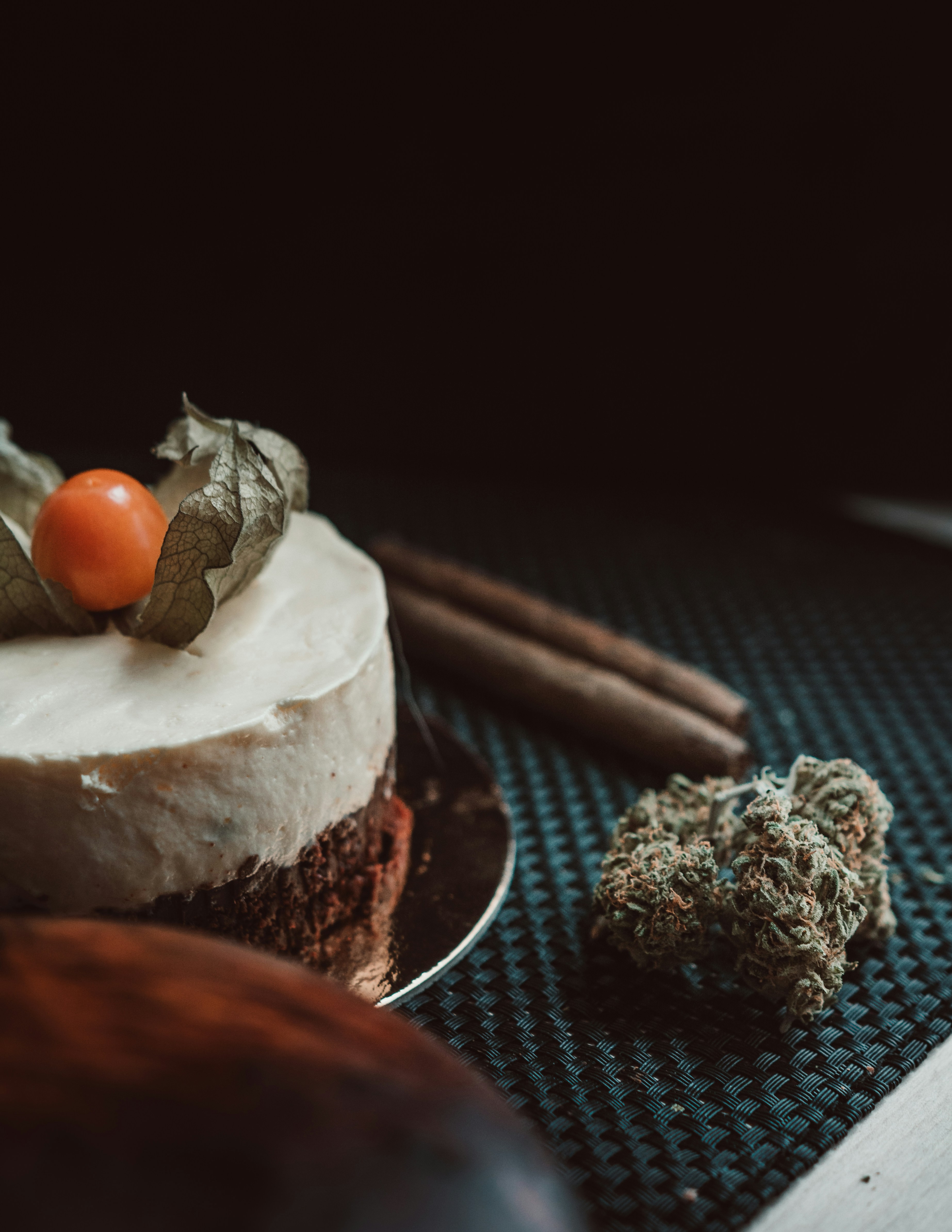 Creamy cake topped with a golden berry and leaves, accompanied by rolled herbs and a textured backdrop.