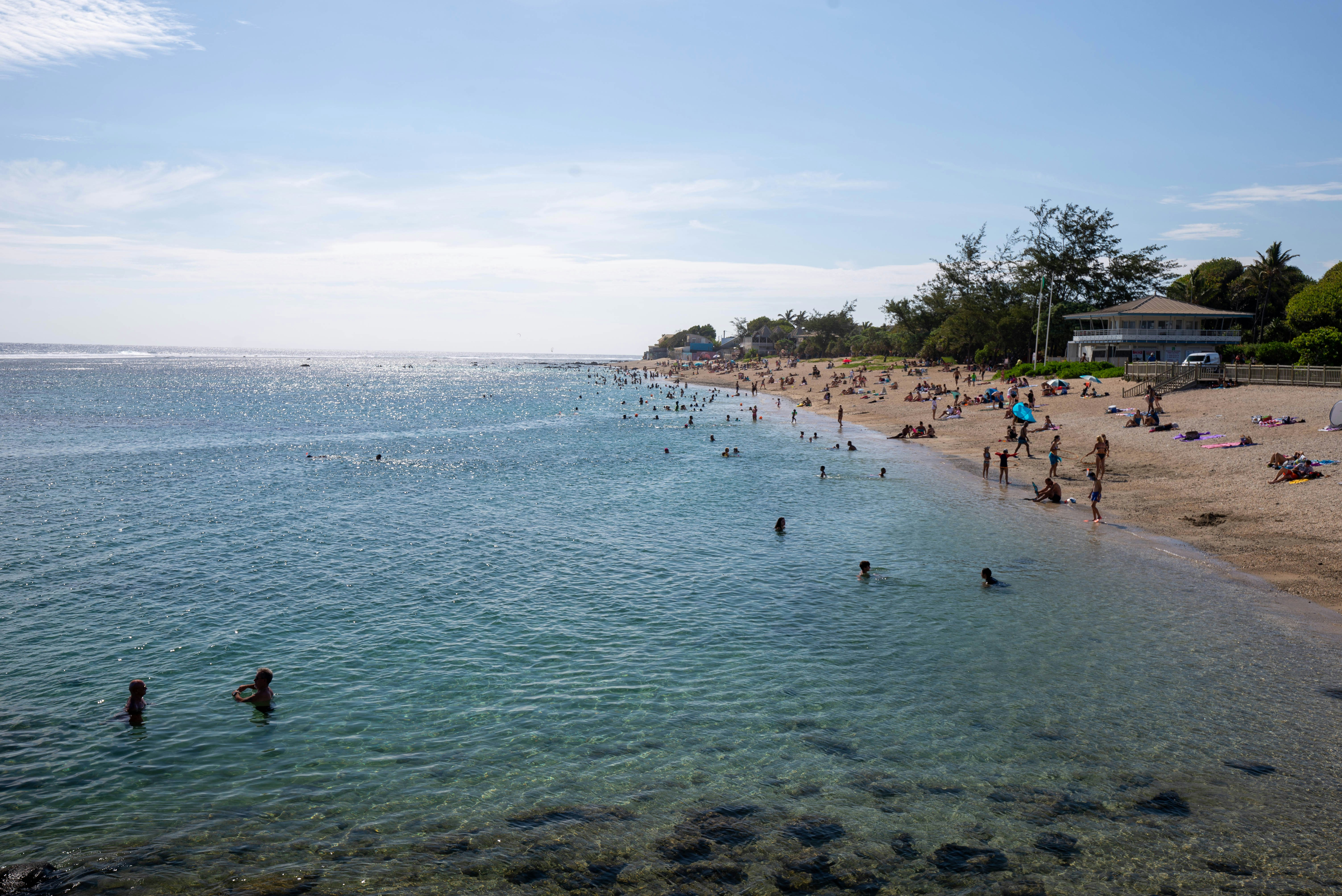 People enjoying a sunny day at a crowded beach with clear blue waters and lush greenery nearby.