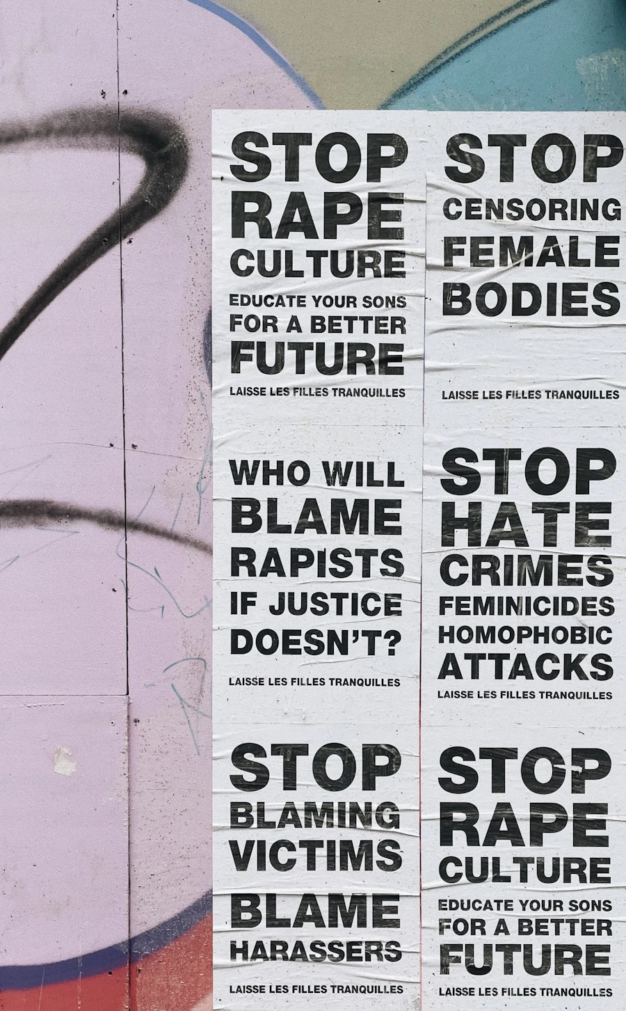 Stacked protest posters call to stop rape culture, victim blaming, and hate crimes against women and marginalized groups. The white flyers are pasted over a pastel graffiti wall.