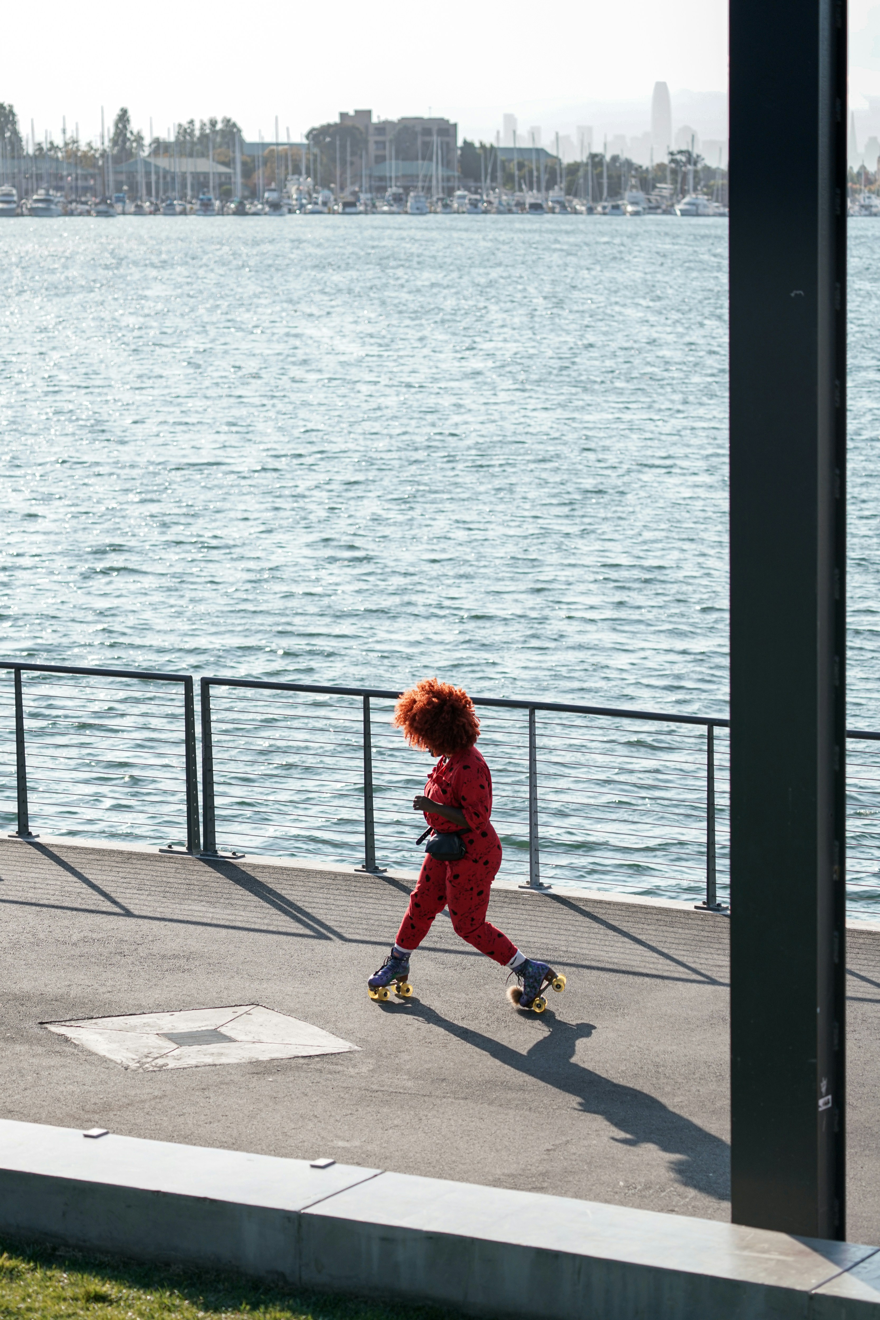 Crimson-clad roller skater glides along a waterfront promenade beside a railing, with a calm harbor and distant city skyline in the background.