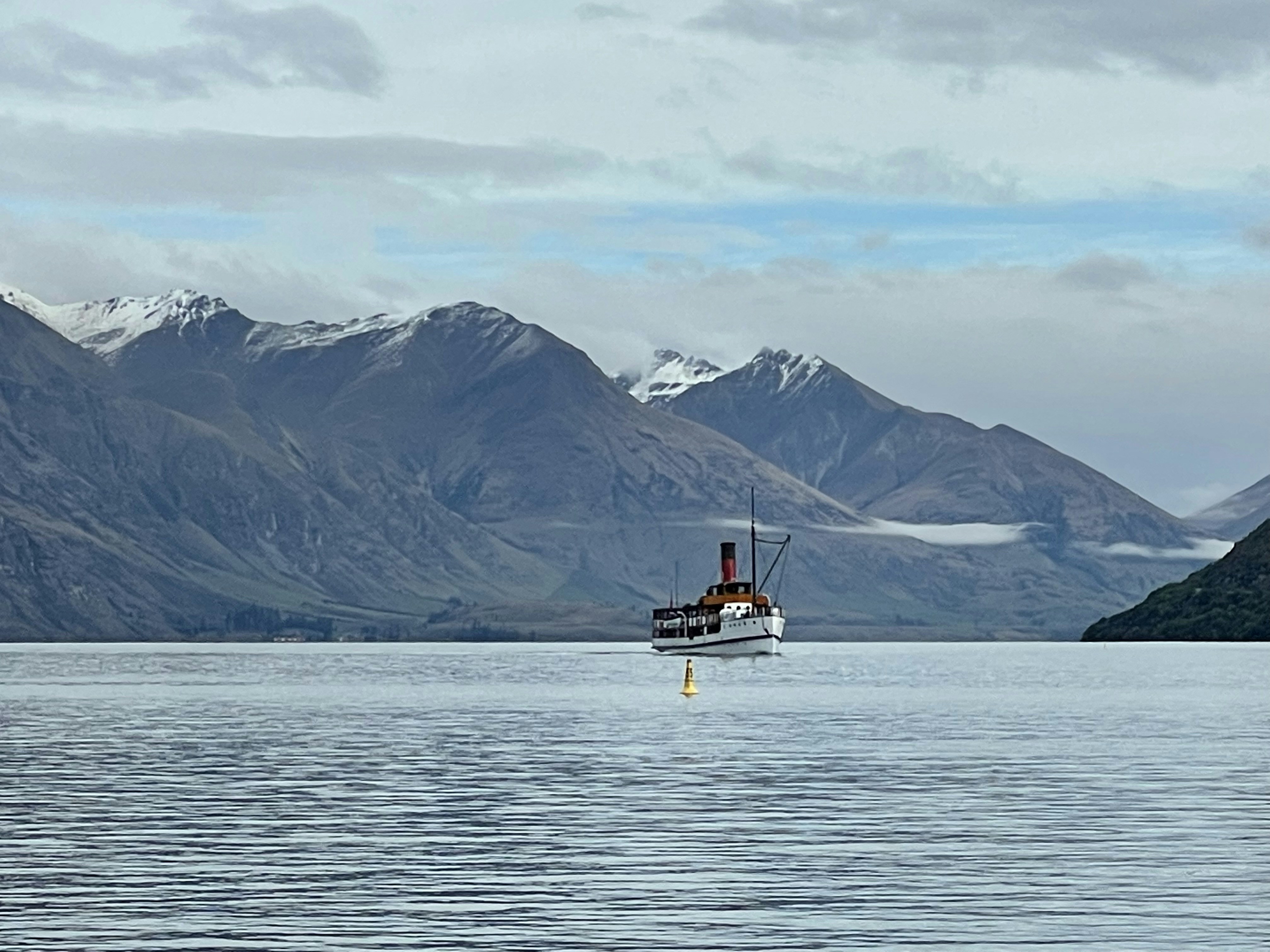 Historic steamboat gliding across a serene lake surrounded by majestic mountains and a hint of snow, showcasing nature's splendor.