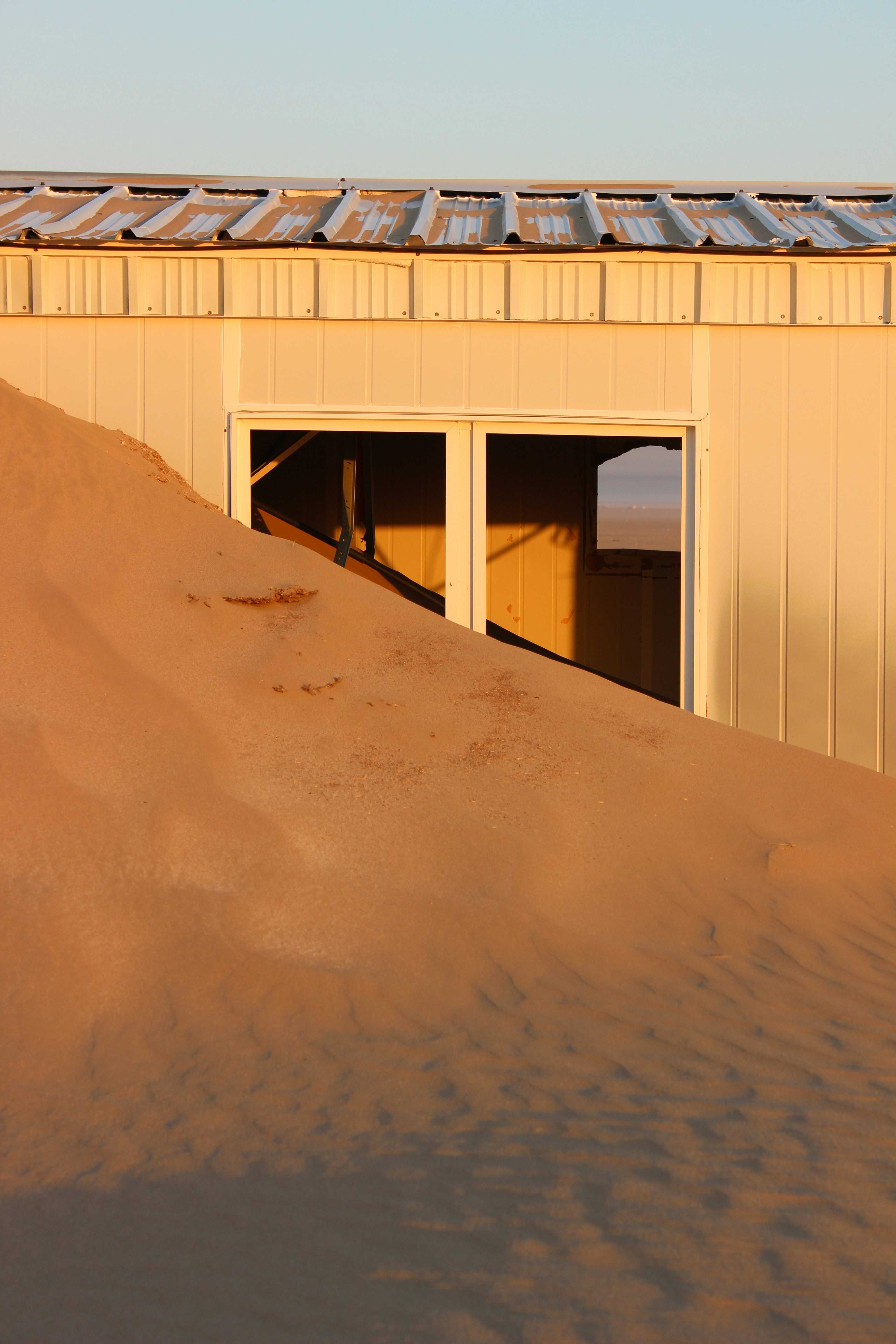 A sunlit, half-buried structure in a desert, with sand drifting against its metallic exterior, revealing a glimpse of the interior.