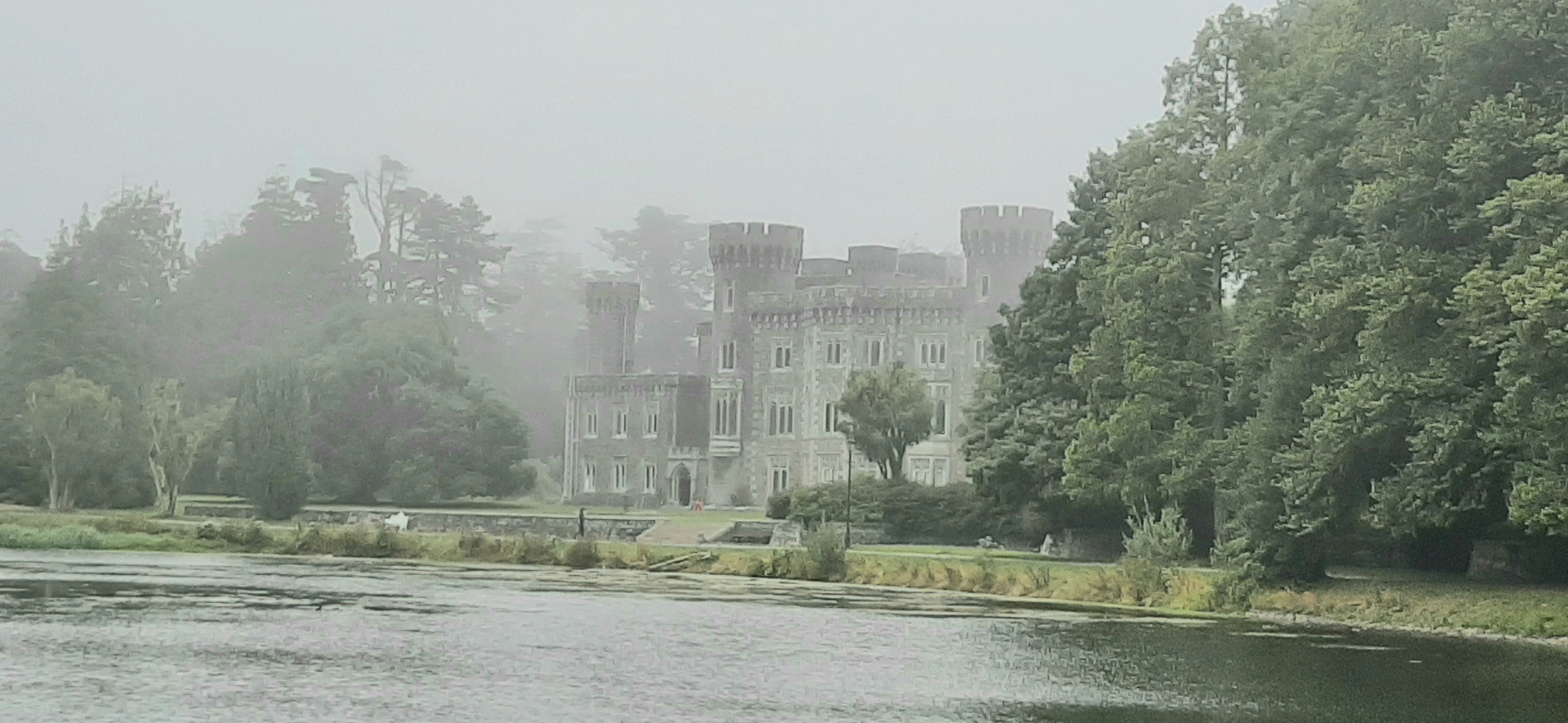 Misty view of a historic castle nestled among lush greenery by a tranquil lake, evoking a sense of mystery and serenity.