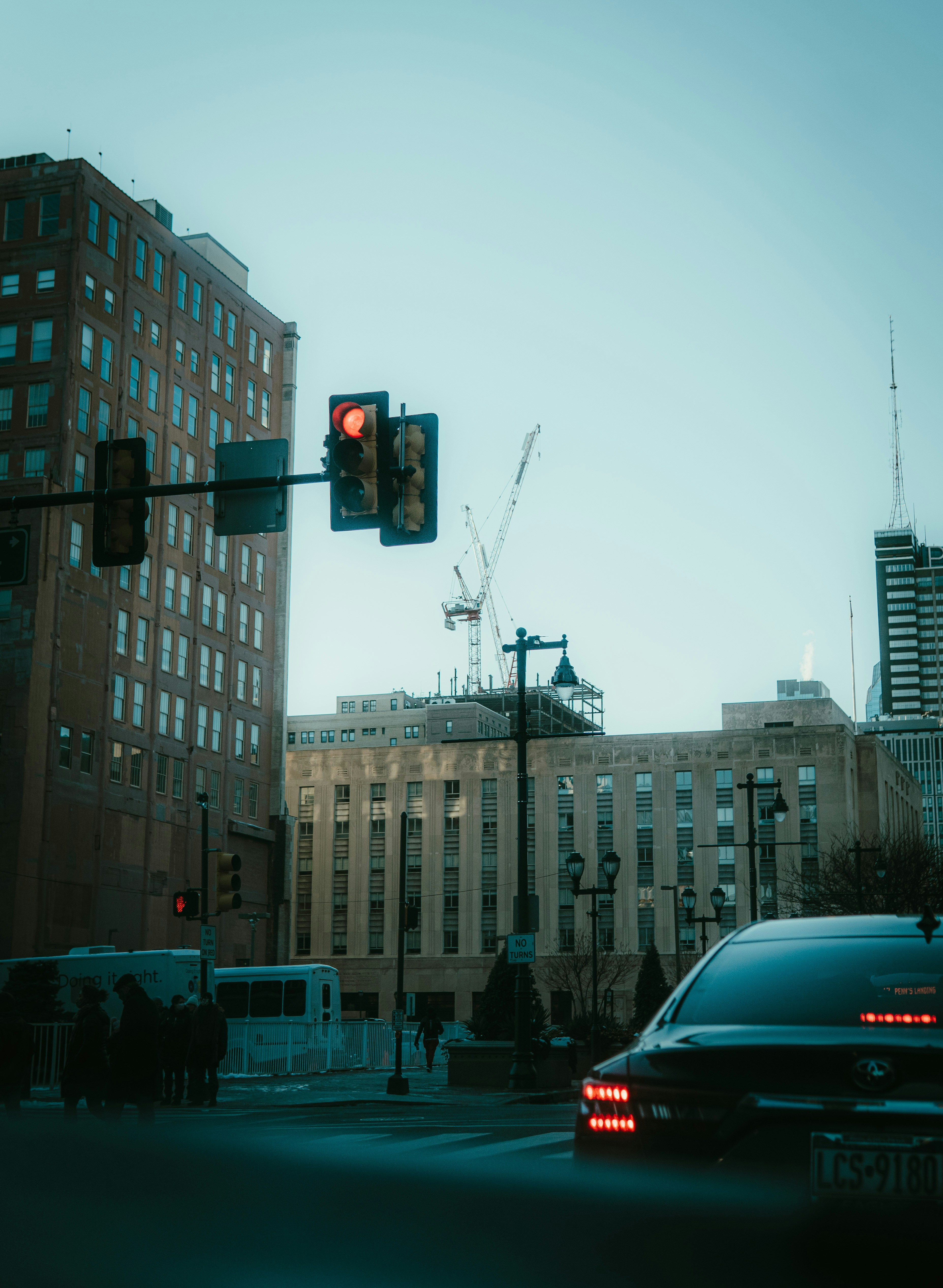 Traffic lights signal a halt at a bustling city intersection, with a backdrop of towering buildings and construction cranes. The scene captures the essence of urban life in motion.