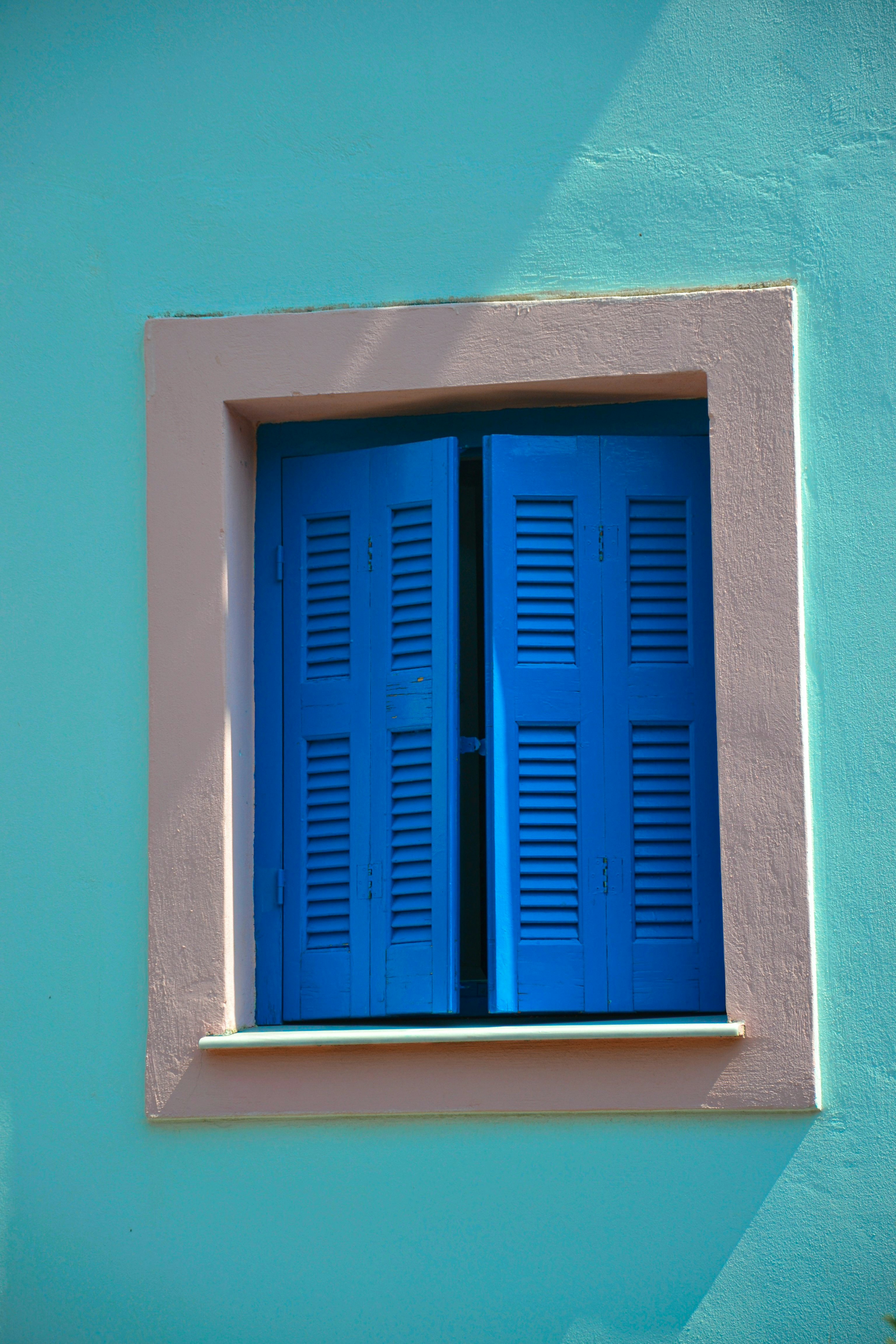Vibrant blue shutters framed by a soft pastel wall, showcasing a coastal architectural style. The interplay of colors adds a refreshing touch to the scene.