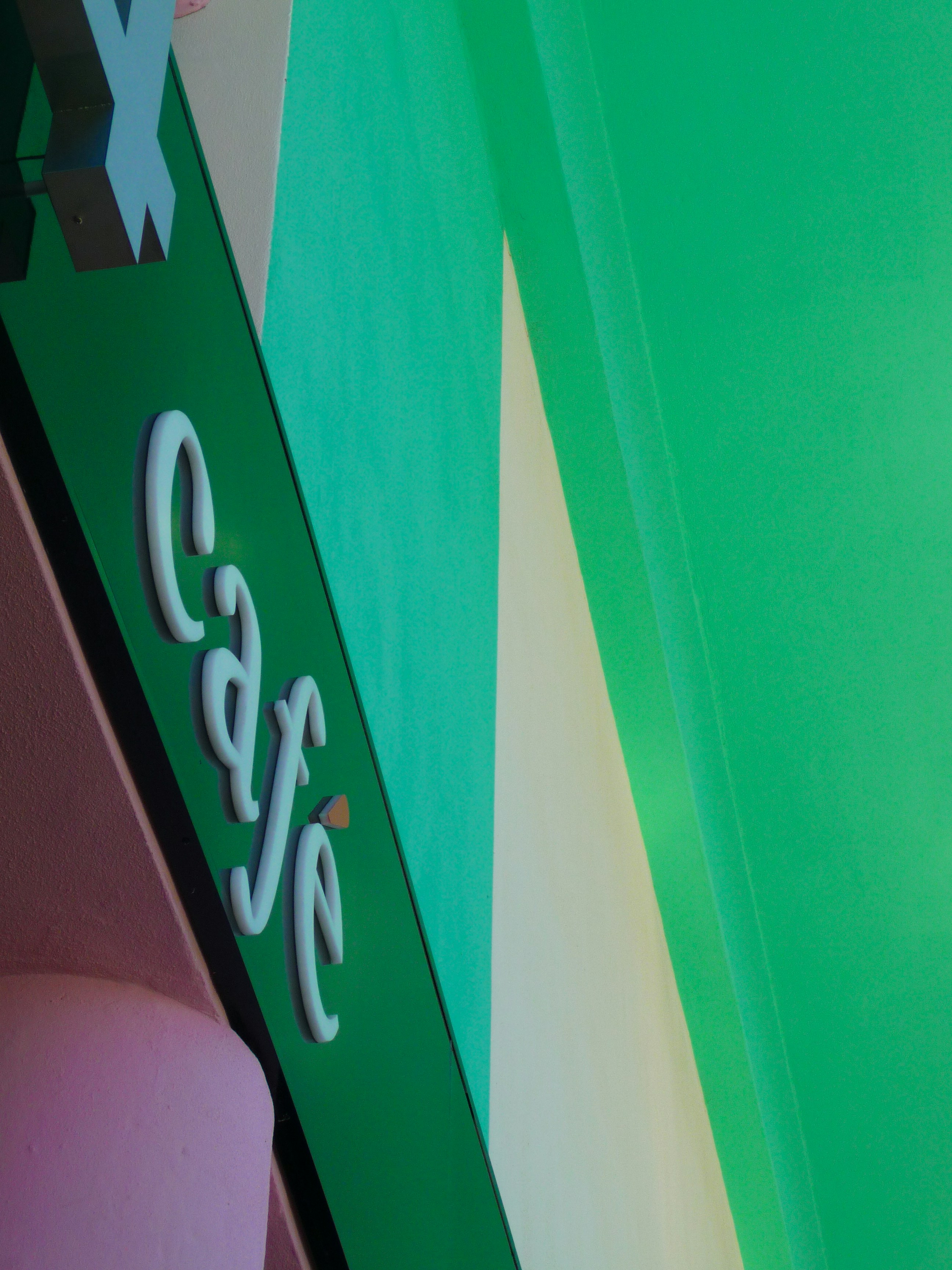 Close-up of a glossy green sign with white cursive letters on a tilted, color-blocked façade; bright green and teal panels create abstract geometry.