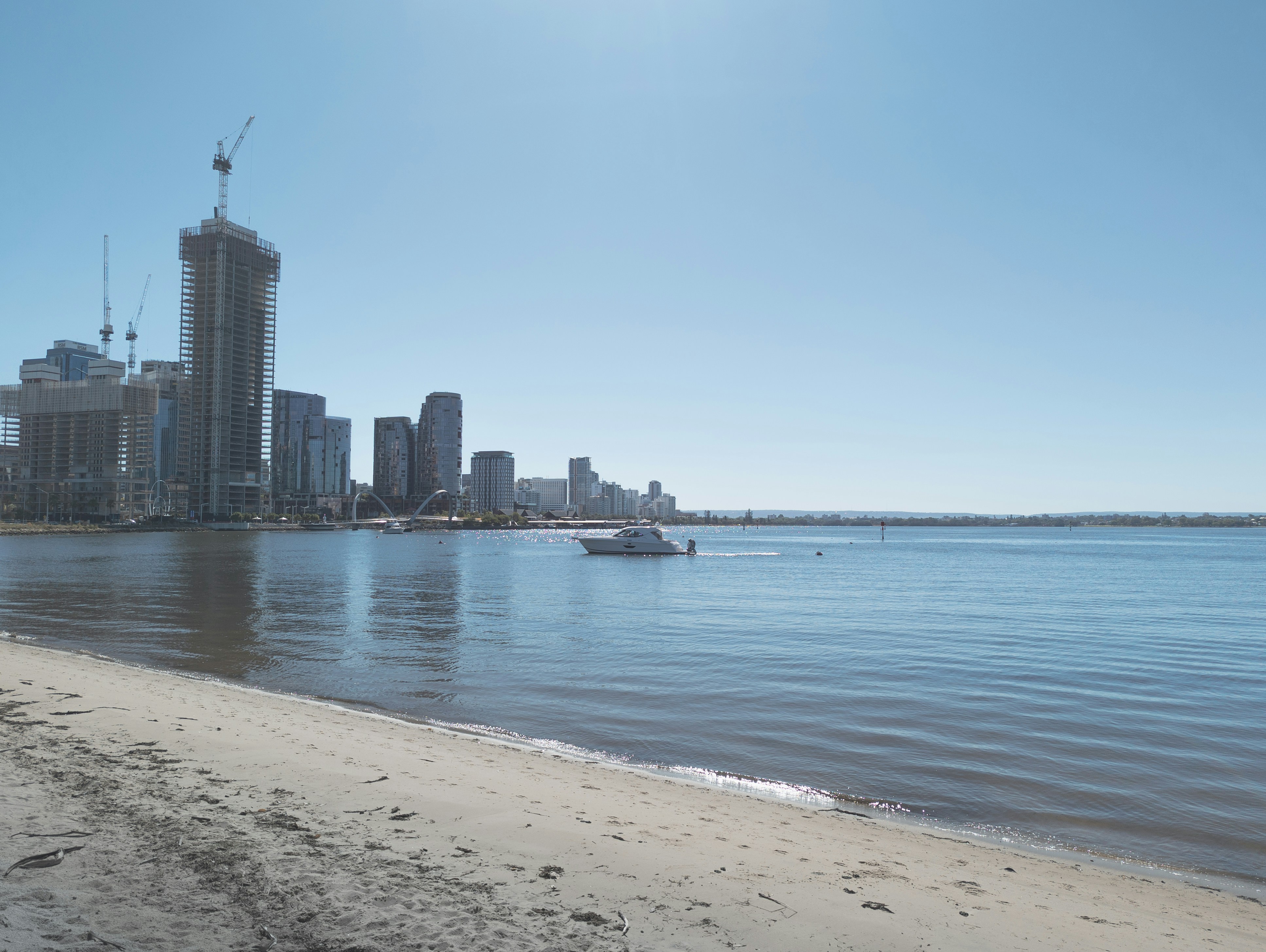 Boat floating near sandy beach with tall city buildings under a clear blue sky.