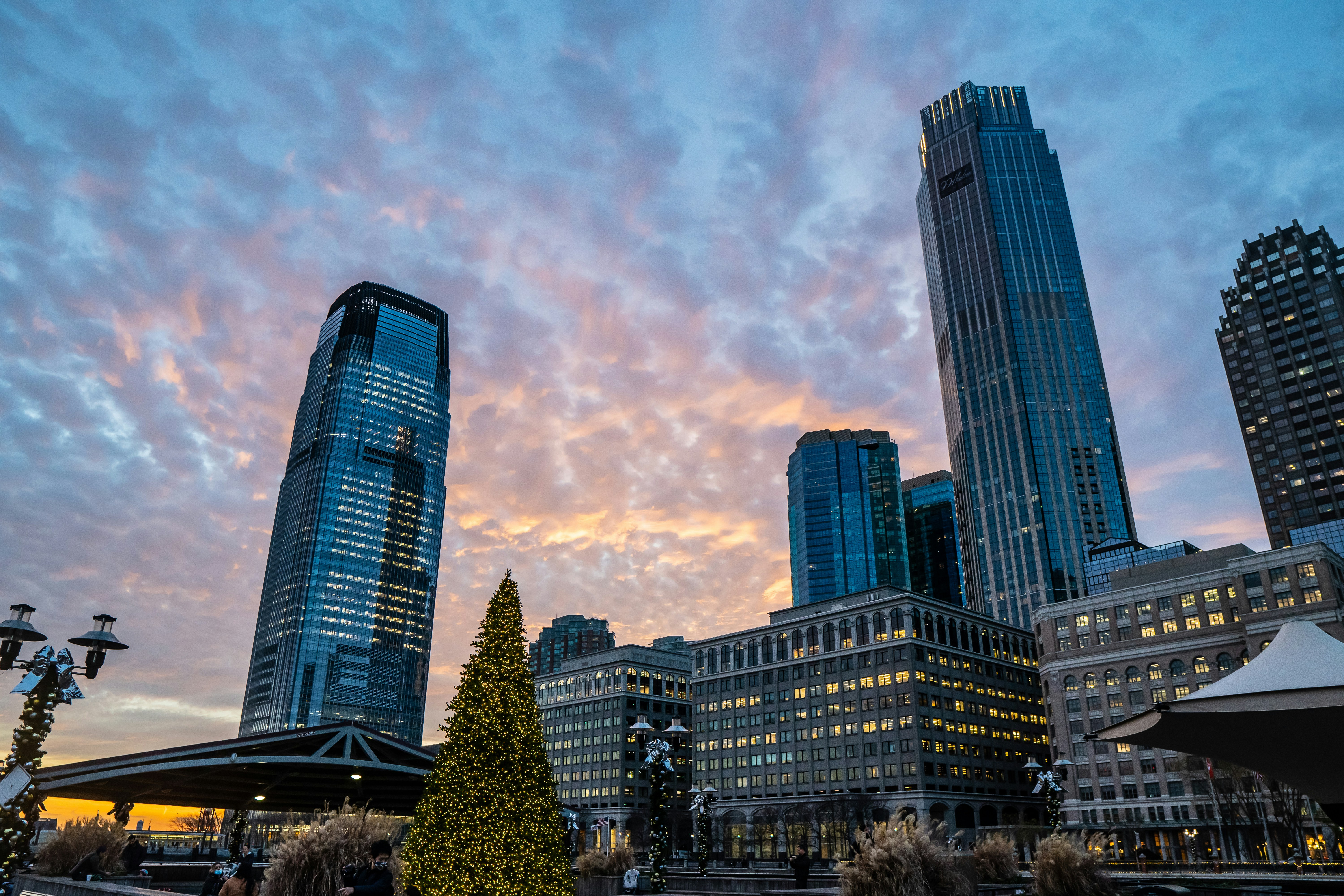Illuminated Christmas tree in a bustling urban plaza, framed by towering skyscrapers under a vibrant sunset sky.