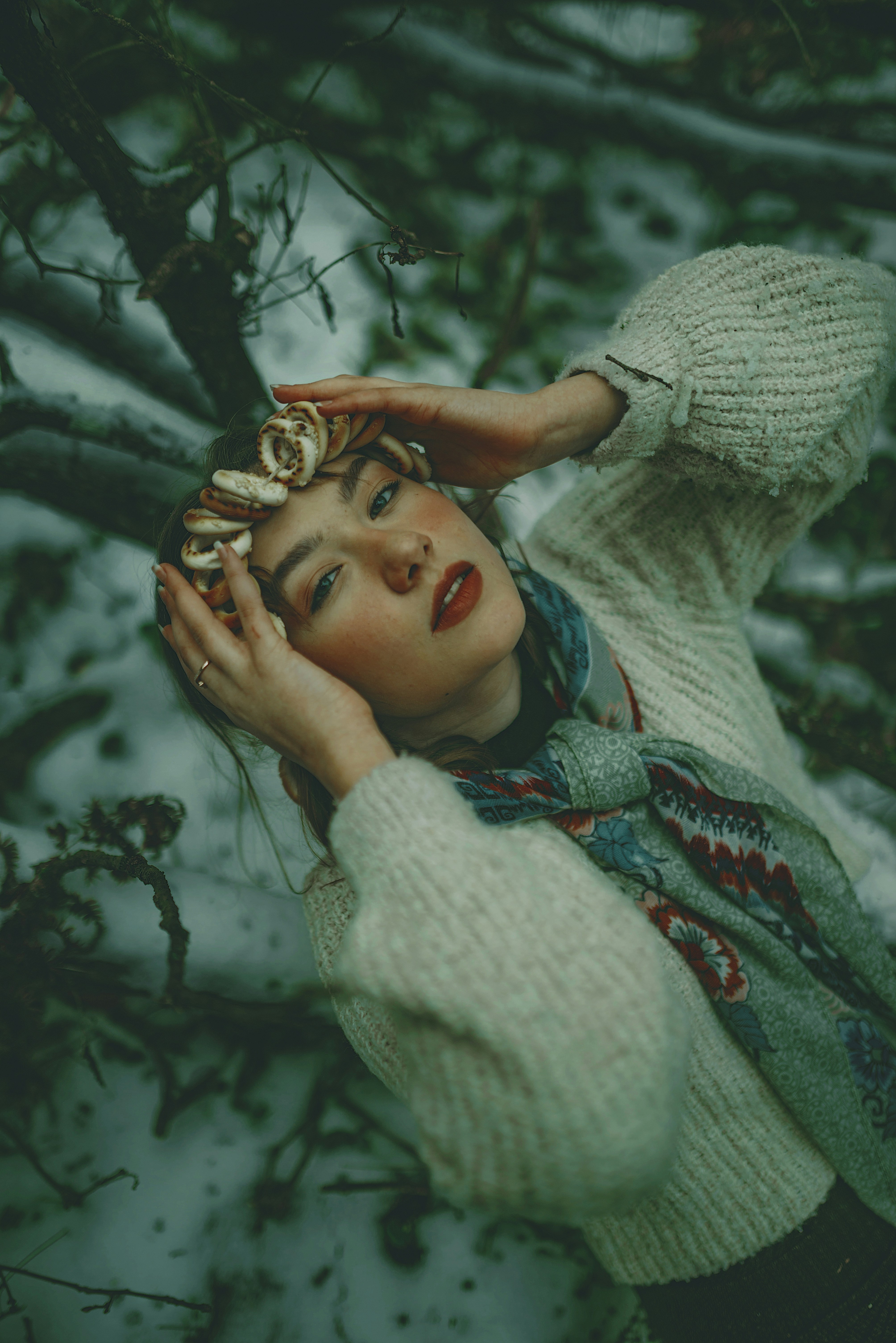 A woman adorned with a delicate crown of mushrooms gazes thoughtfully, surrounded by branches in a snowy landscape.