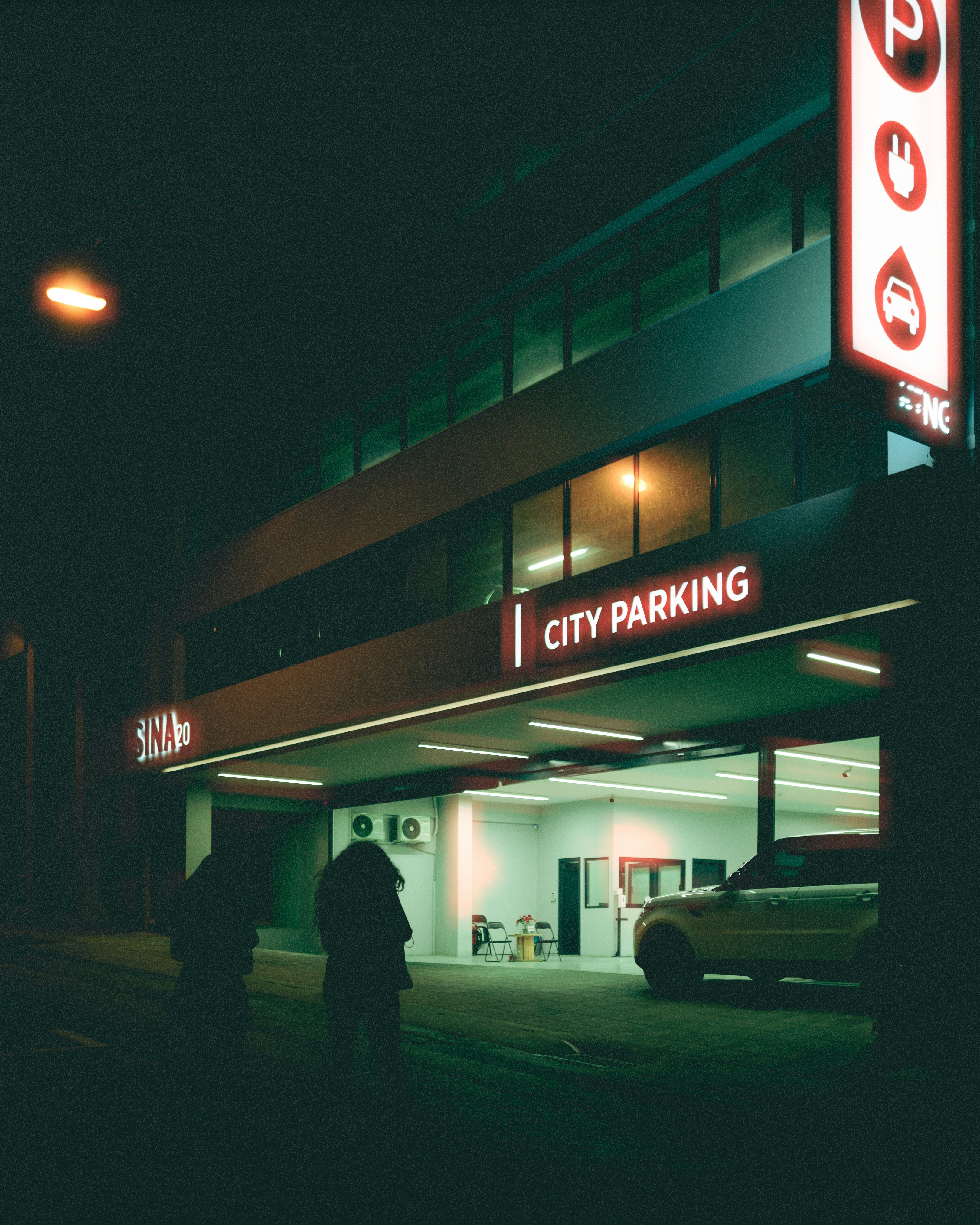 A modern city parking facility illuminated at night, showcasing its sleek architecture and vibrant signage. The interior is visible, hinting at a welcoming atmosphere.
