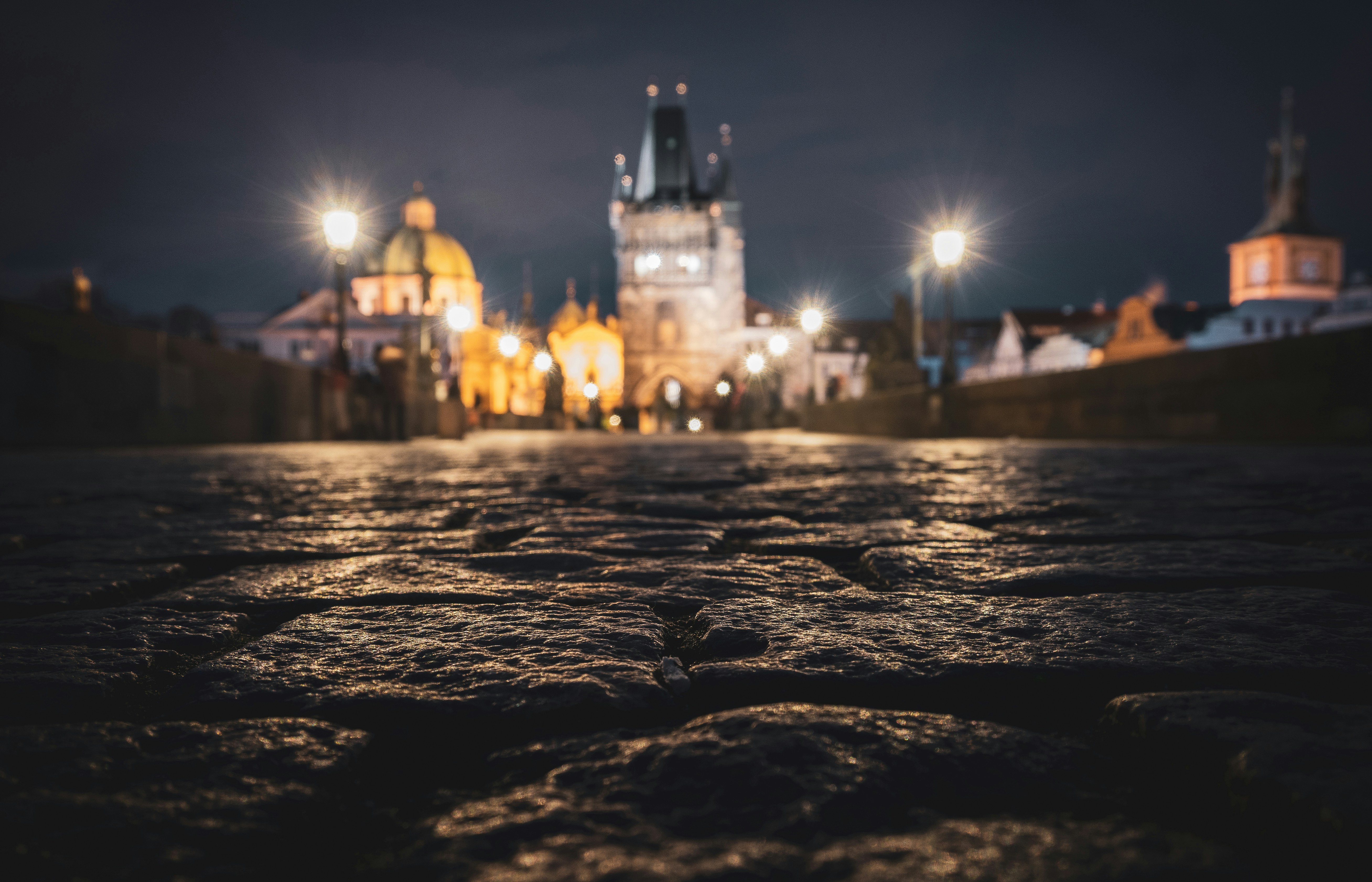 Cobblestone street illuminated by street lamps, leading towards a historic tower in a quaint town at night.