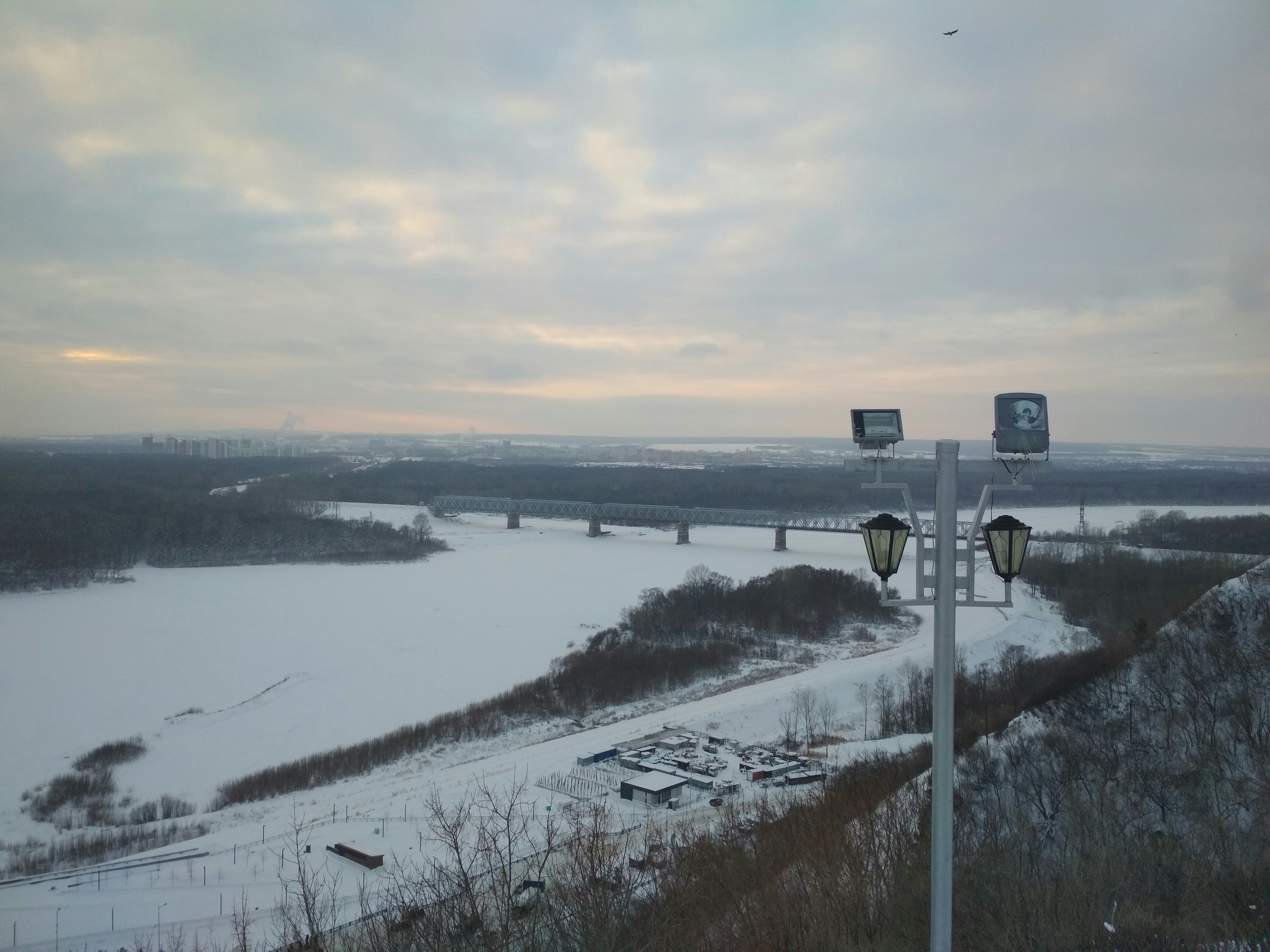 Winter landscape photograph of a snow-covered river valley with a lamppost in the foreground and a distant bridge under an overcast sky.