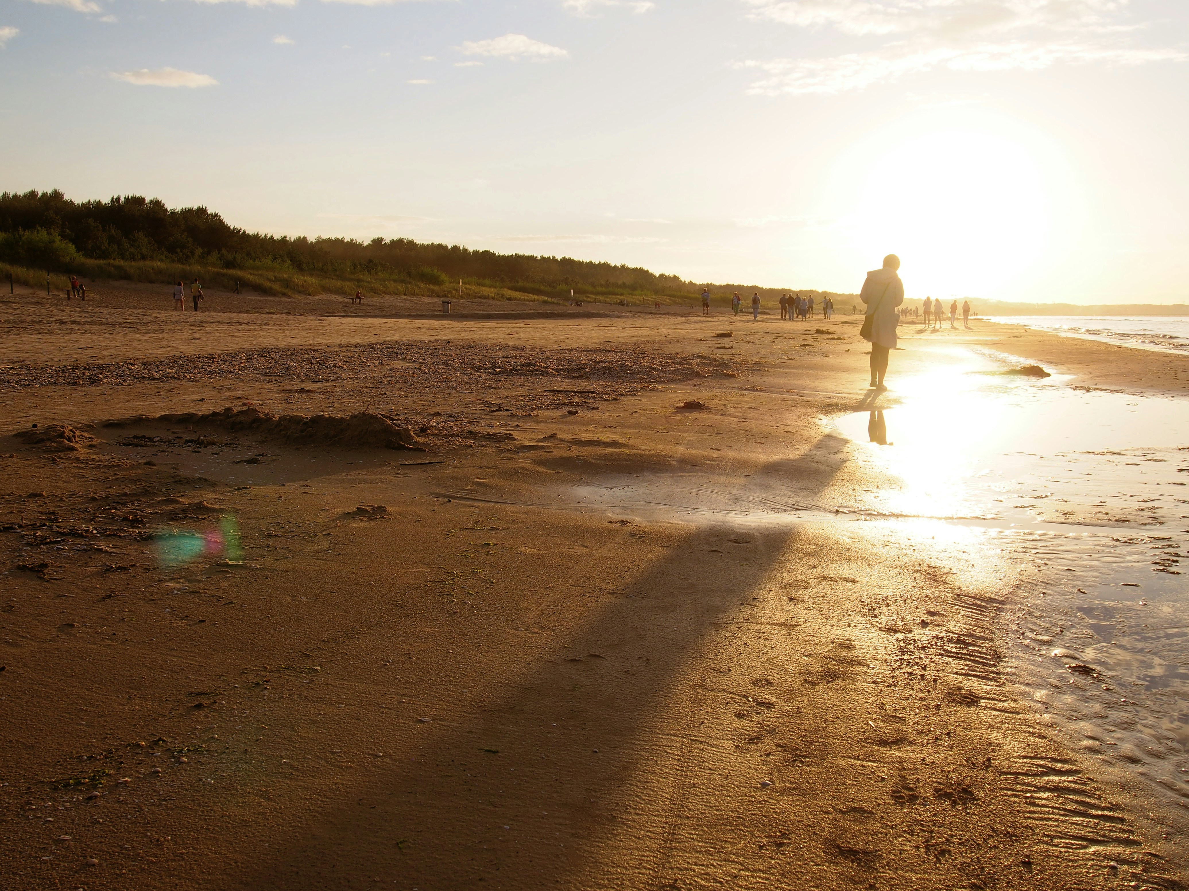 Silhouette of a person walking along a sunlit beach with gentle waves and a distant horizon.