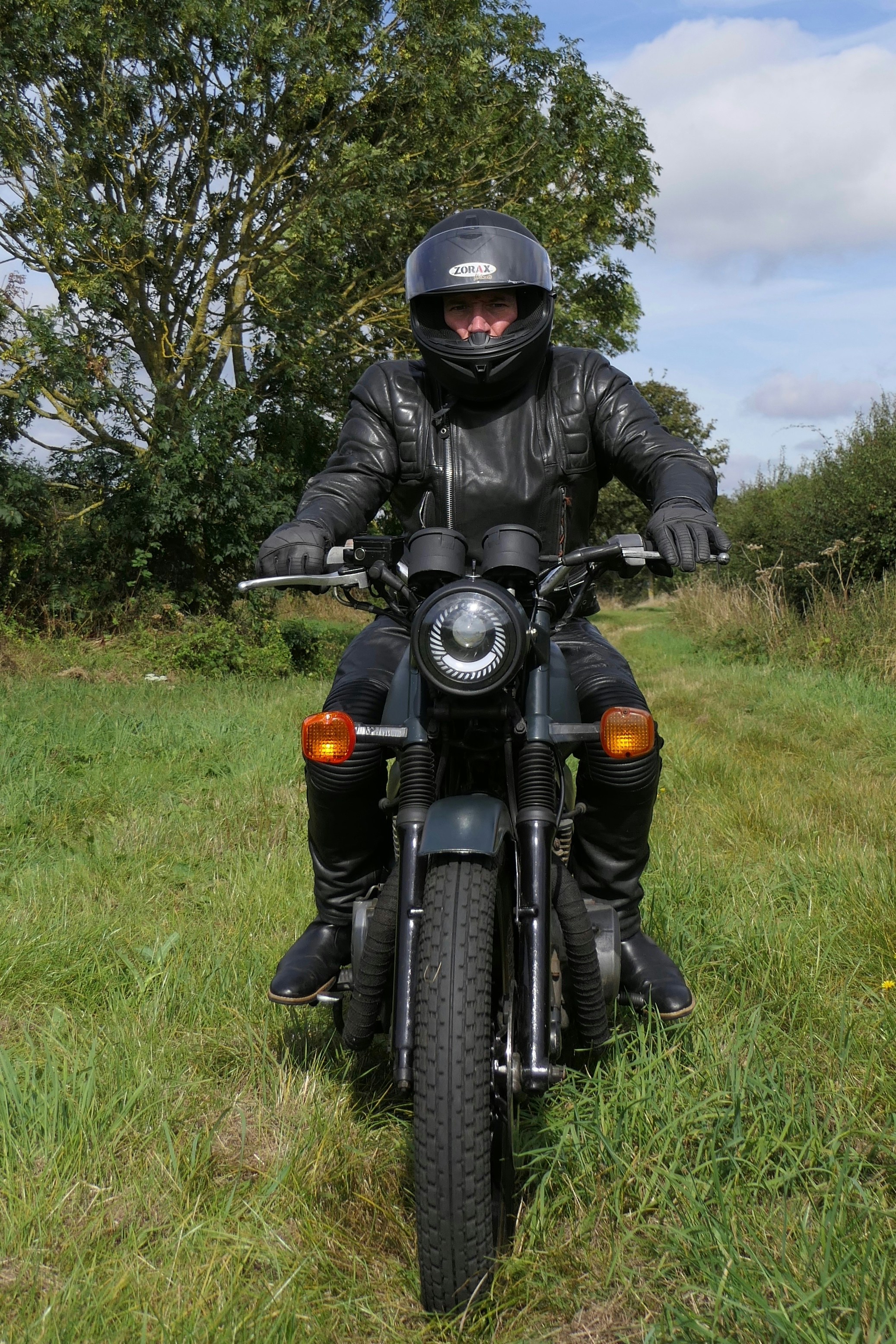 A motorcyclist in black leather sits astride a motorcycle, facing the camera in a grassy field with trees and a blue sky.