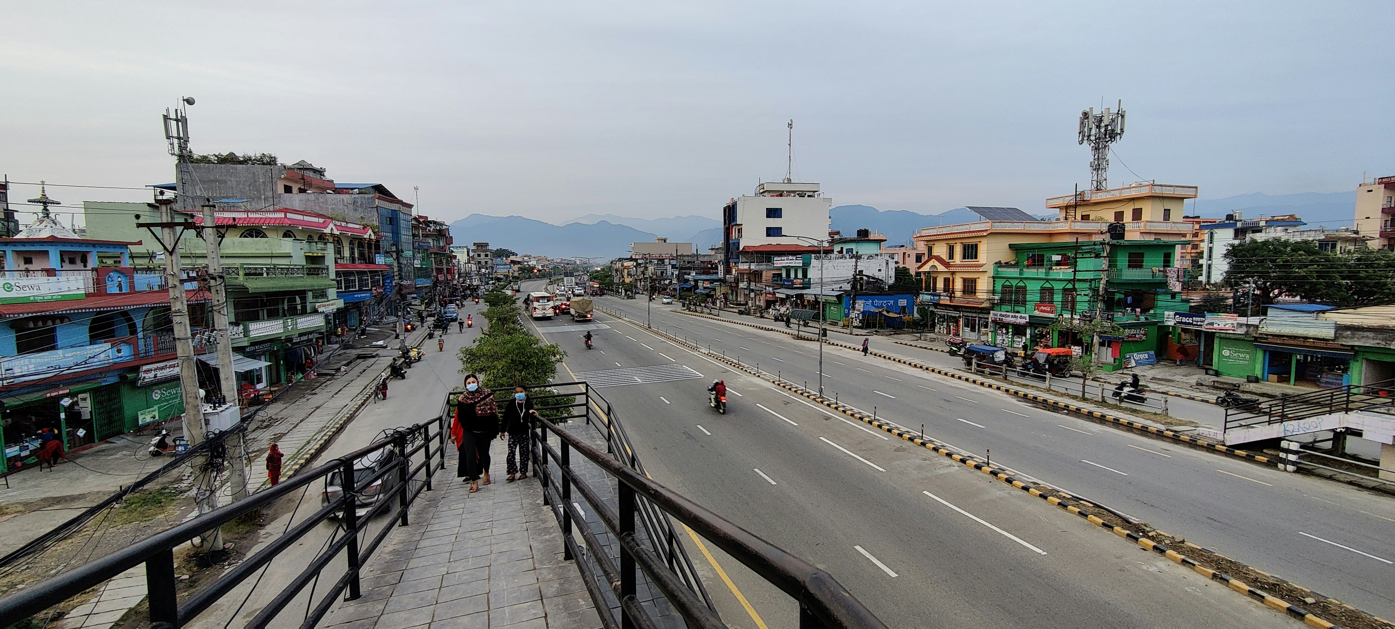Quiet city street view from a pedestrian bridge with distant mountains under a cloudy sky.