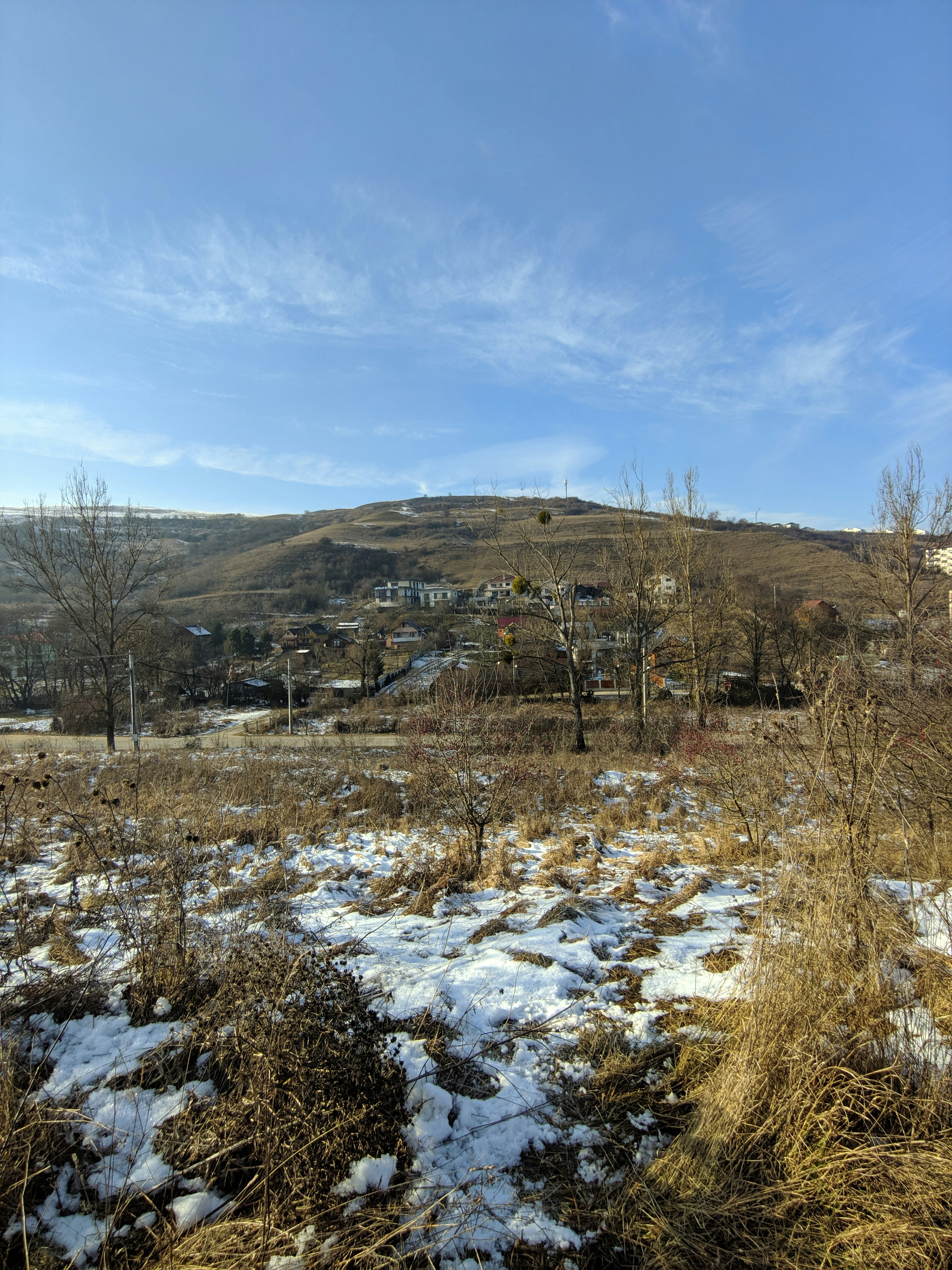 Barren hillside with patches of snow and sparse vegetation under a clear blue sky, revealing a quiet residential area in the valley below.