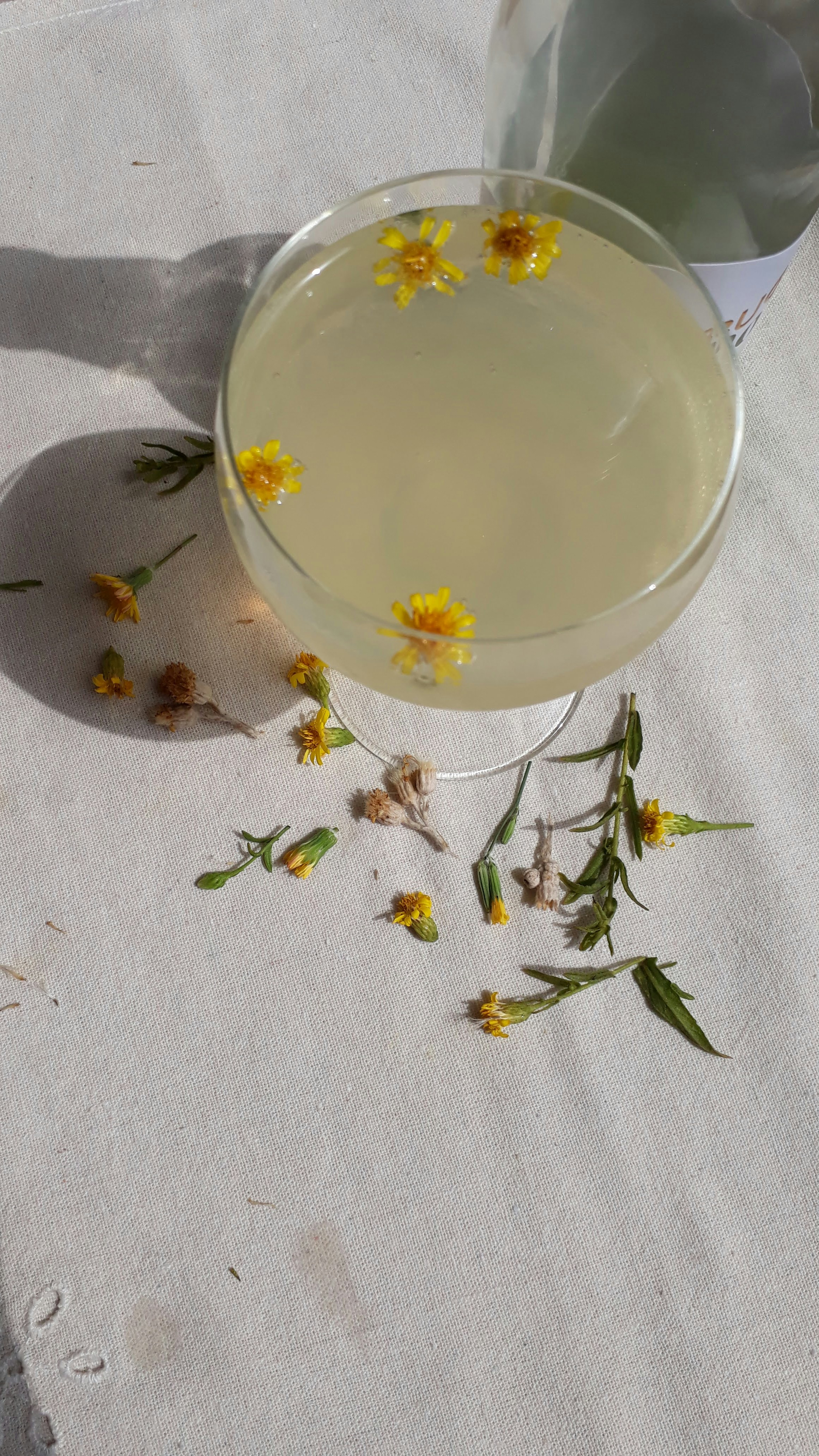 A clear cocktail glass filled with a light beverage, adorned with yellow flowers, set against a textured fabric background. A bottle partially visible in the background adds context.