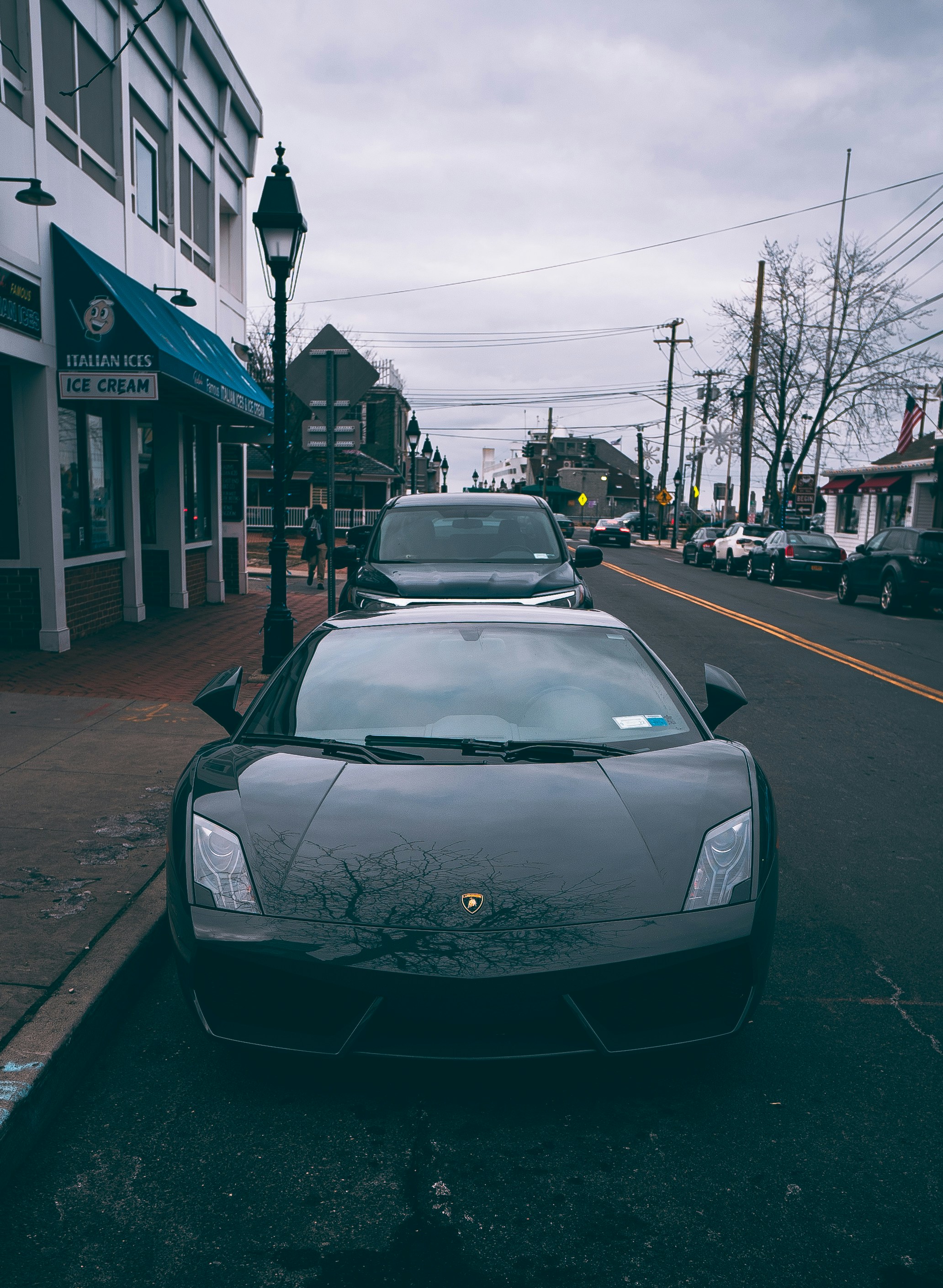 Sleek black Lamborghini parked on a city street, surrounded by urban architecture and a cloudy sky. The scene captures the contrast between luxury and everyday life.