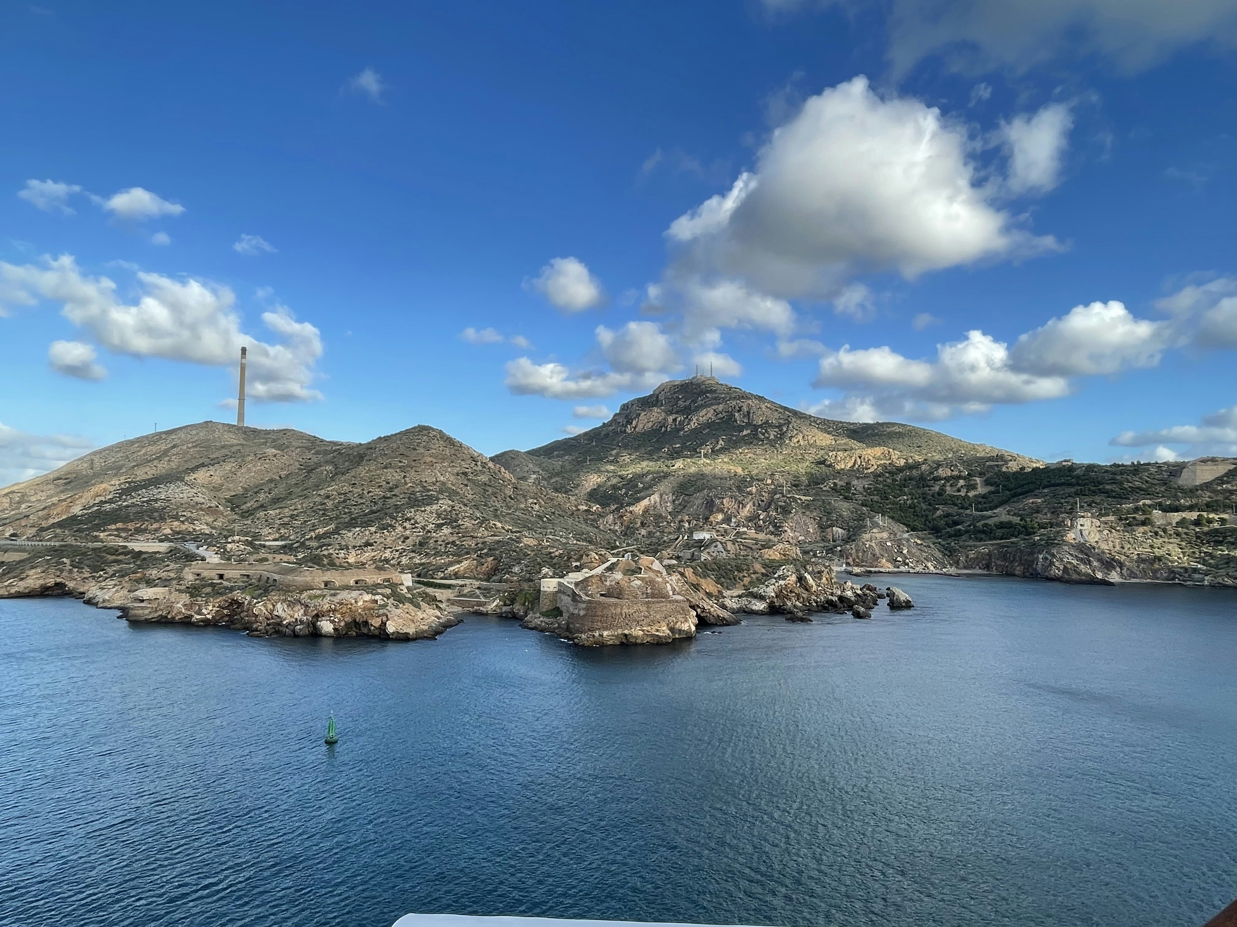 Expansive view of rugged hills and blue sea under a sky dotted with fluffy clouds.