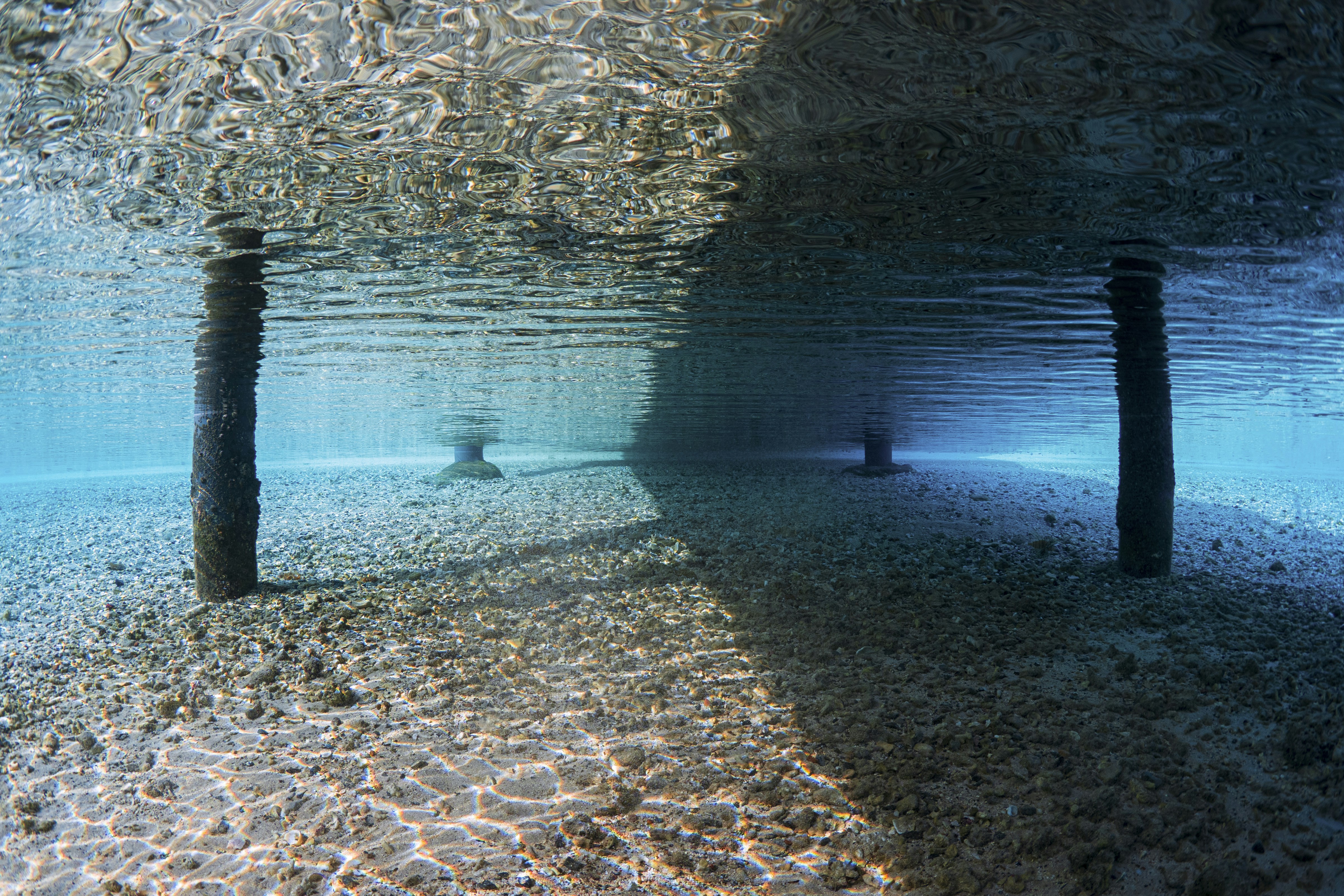 Underwater view showcasing wooden pillars submerged in crystal-clear water, with sunlight creating intricate patterns on the sandy bottom.