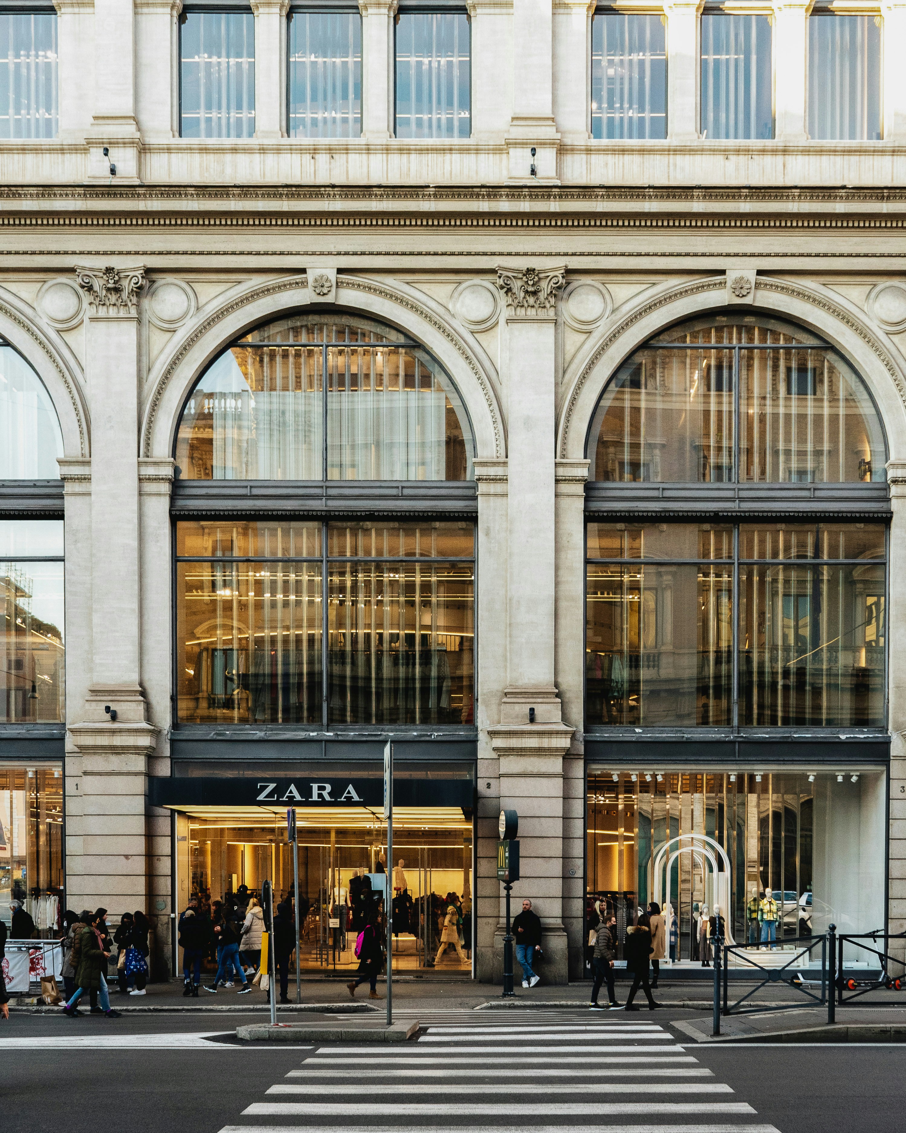 Facade of a Zara store featuring large arched windows and elegant architectural details, bustling with shoppers crossing the street.