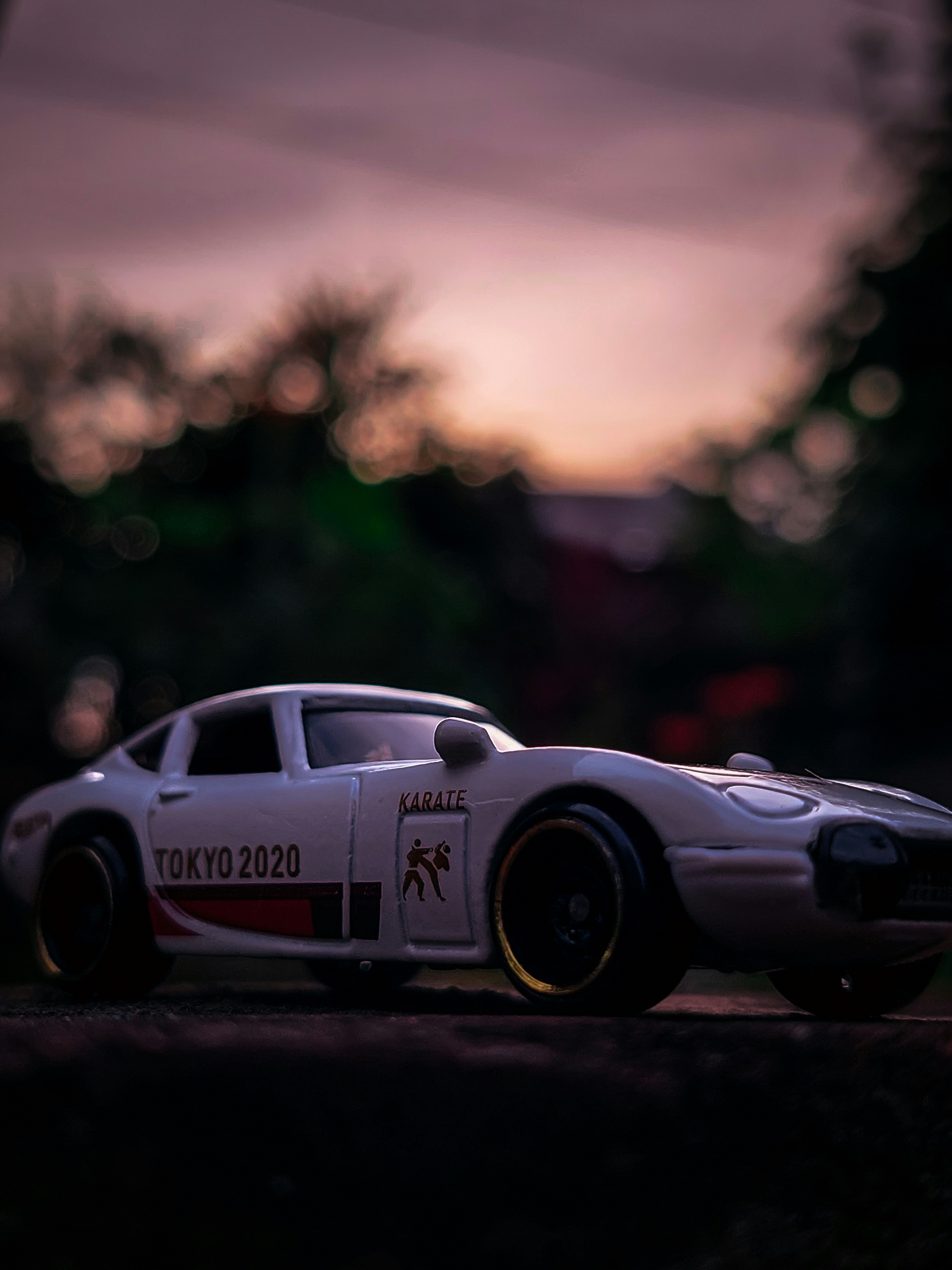 A toy car adorned with 'TOKYO 2020' and a karate motif, set against a vibrant dusk backdrop.