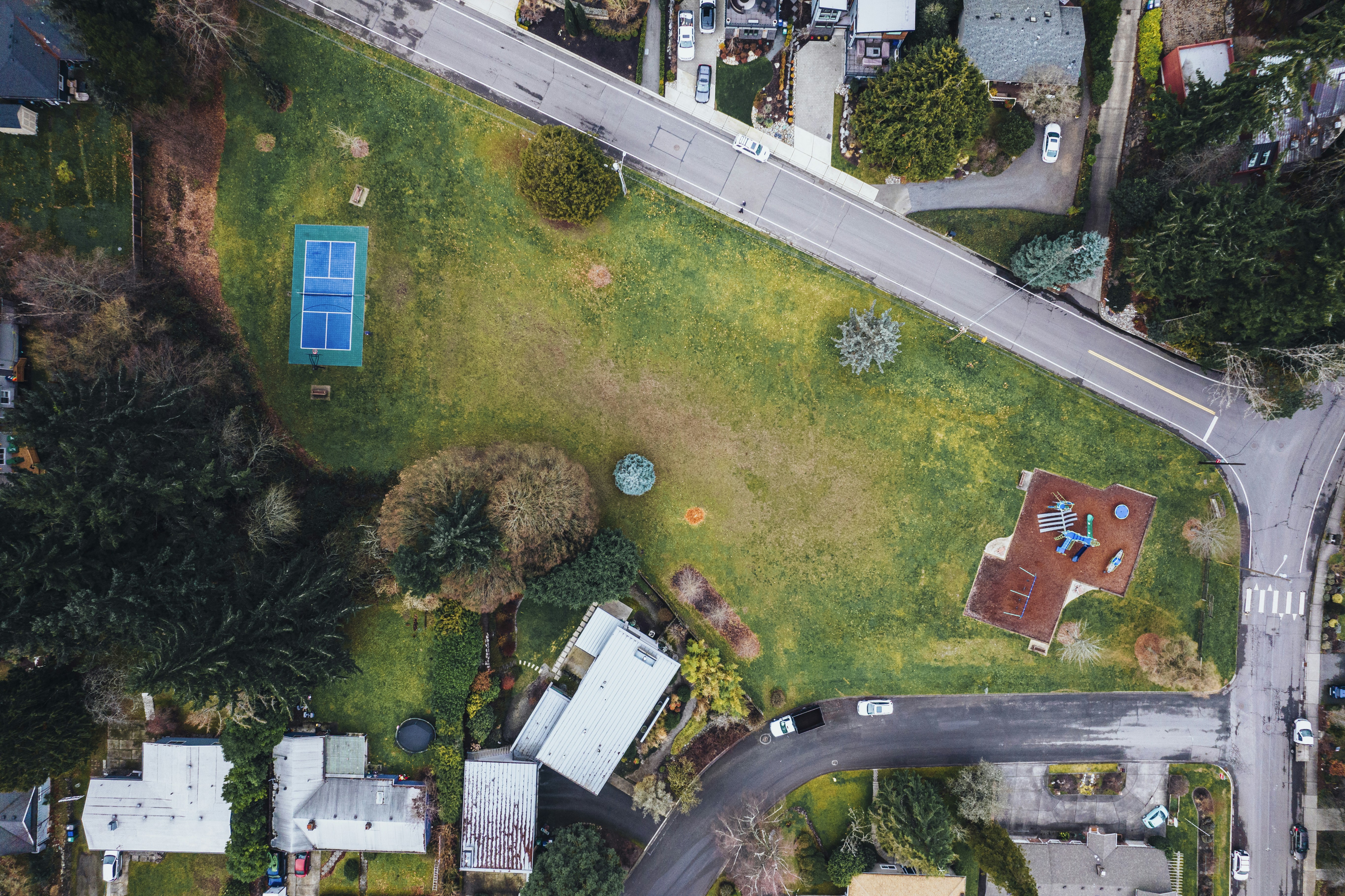 Aerial view of a park featuring a blue tennis court, surrounded by lush greenery and residential areas. The scene captures the blend of urban life and nature.
