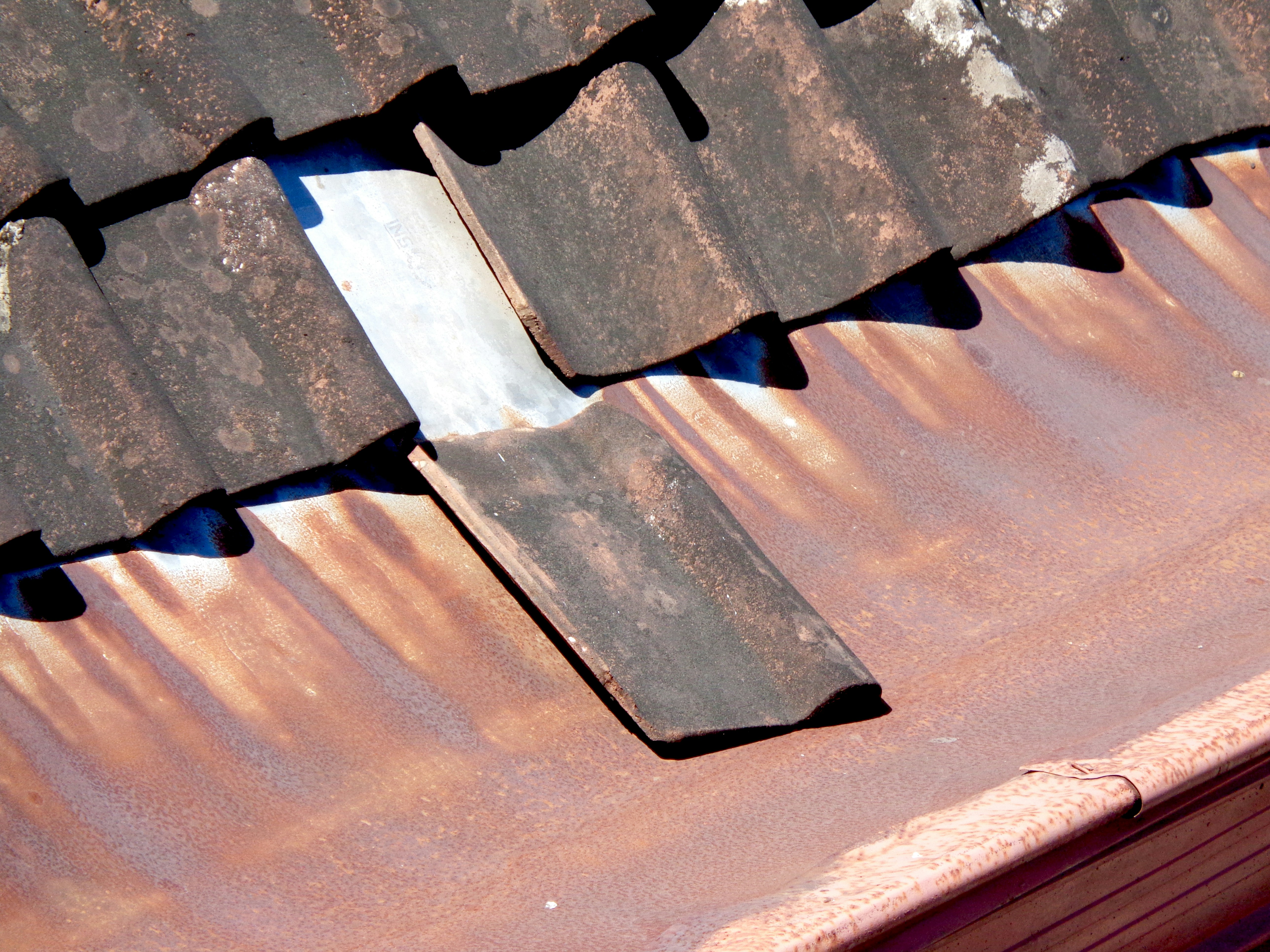 Close-up of a weathered metal roof showcasing a mix of rusted tiles and a contrasting patch of metal. The image highlights the interplay of textures and colors.