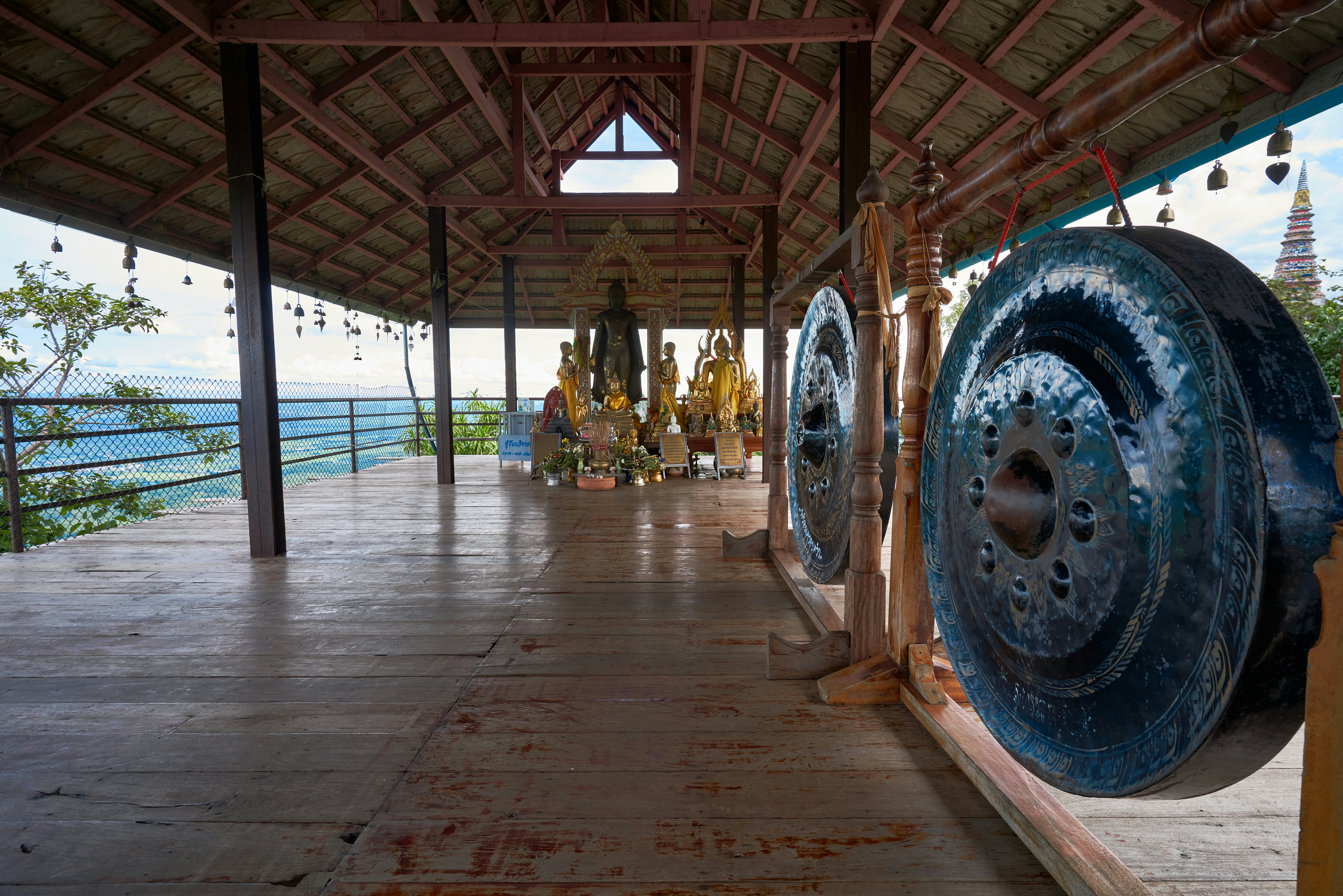 Traditional gongs hang beneath a wooden pavilion overlooking a scenic landscape with statues in the distance.