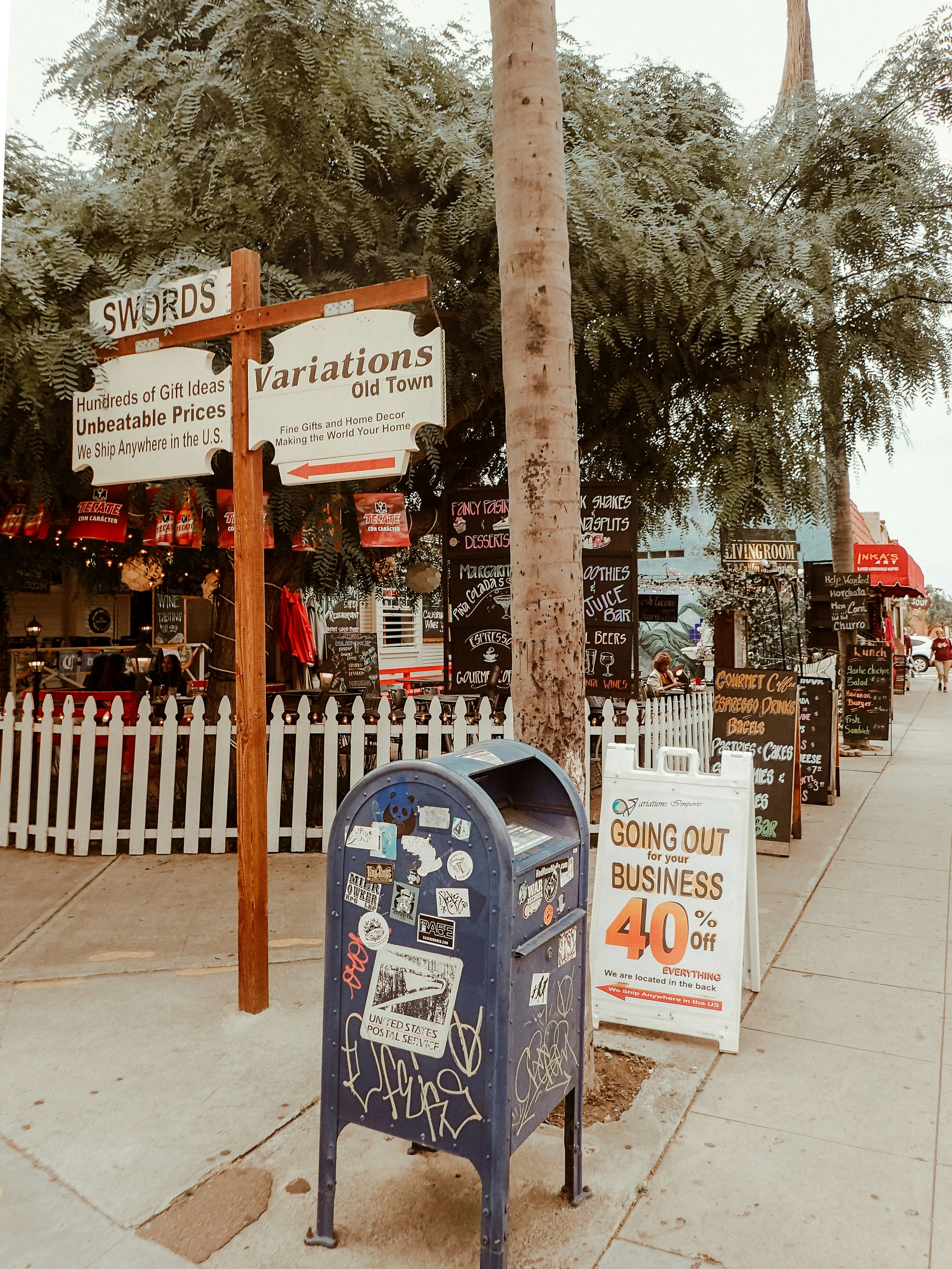 Colorful storefronts and signage line a bustling street, featuring a blue mailbox adorned with stickers. A sign announces a business closing sale.