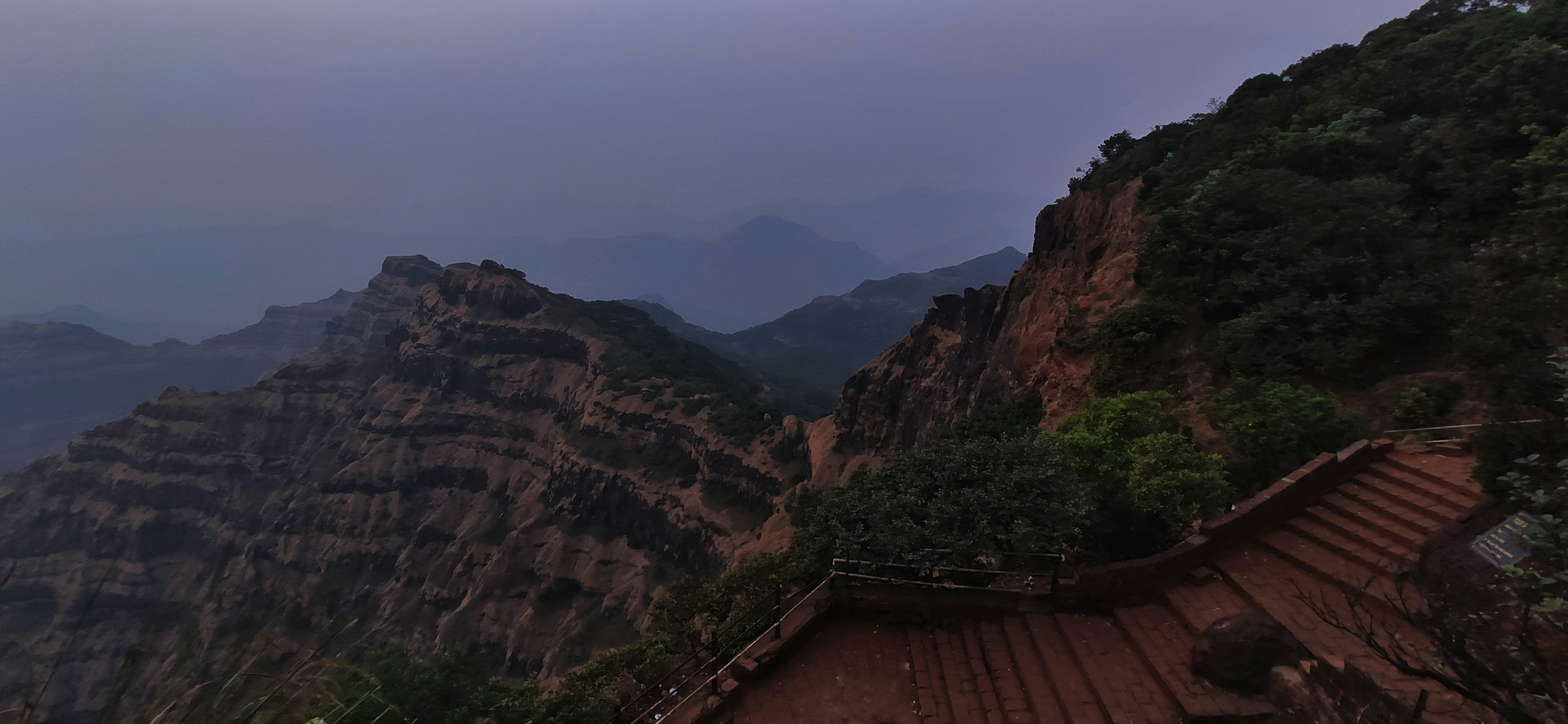 Cliffside path with red-tiled railing winds along a rugged ridge as distant peaks fade into morning mist.