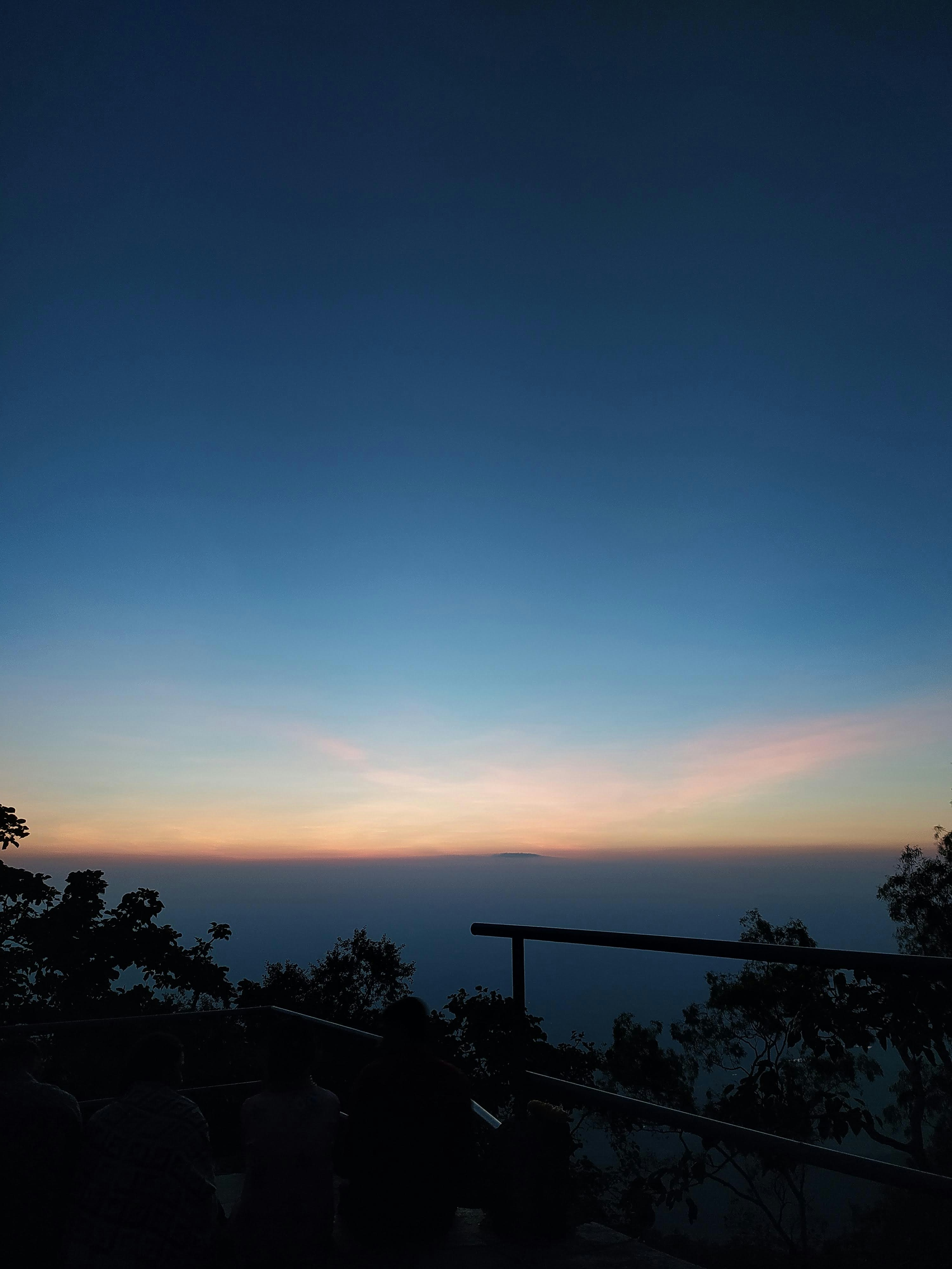 Dusk over a calm horizon with silhouetted railing and trees foregrounding a soft gradient sky; a tranquil viewpoint shown from a cliffside observation area.
