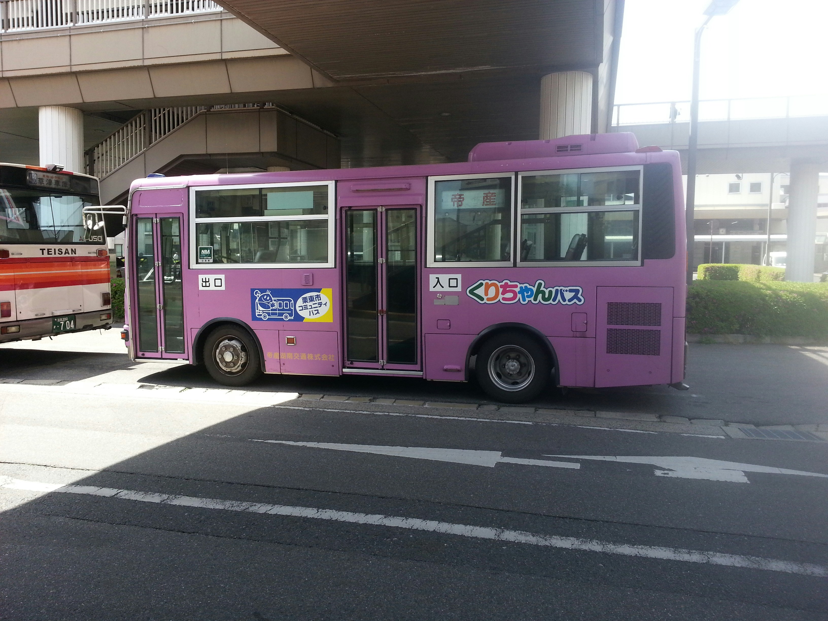 A vibrant purple bus parked beside a traditional vehicle, showcasing its unique design and branding. The setting features urban architecture and greenery in the background.
