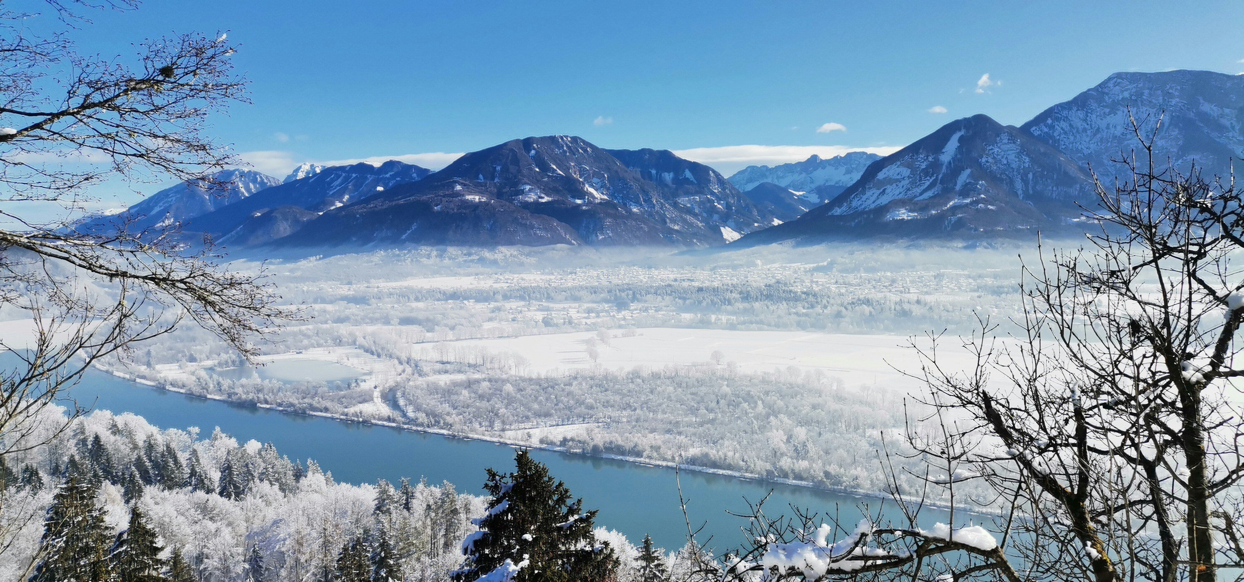 Snow-covered landscape with a winding river and majestic mountains under a clear blue sky. Frosty trees frame the scene beautifully.