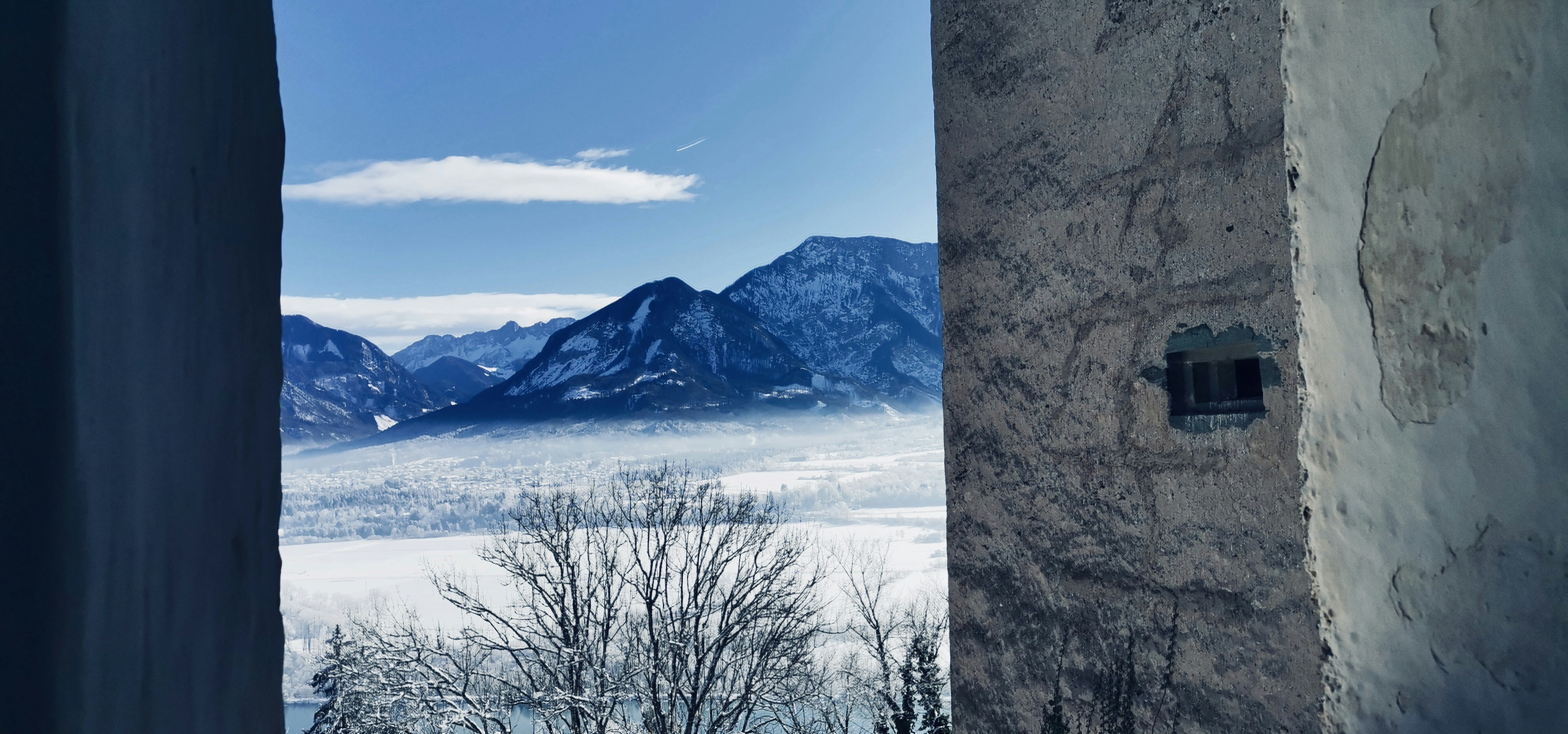 Snow-covered mountains viewed through a rustic stone window under a clear blue sky.