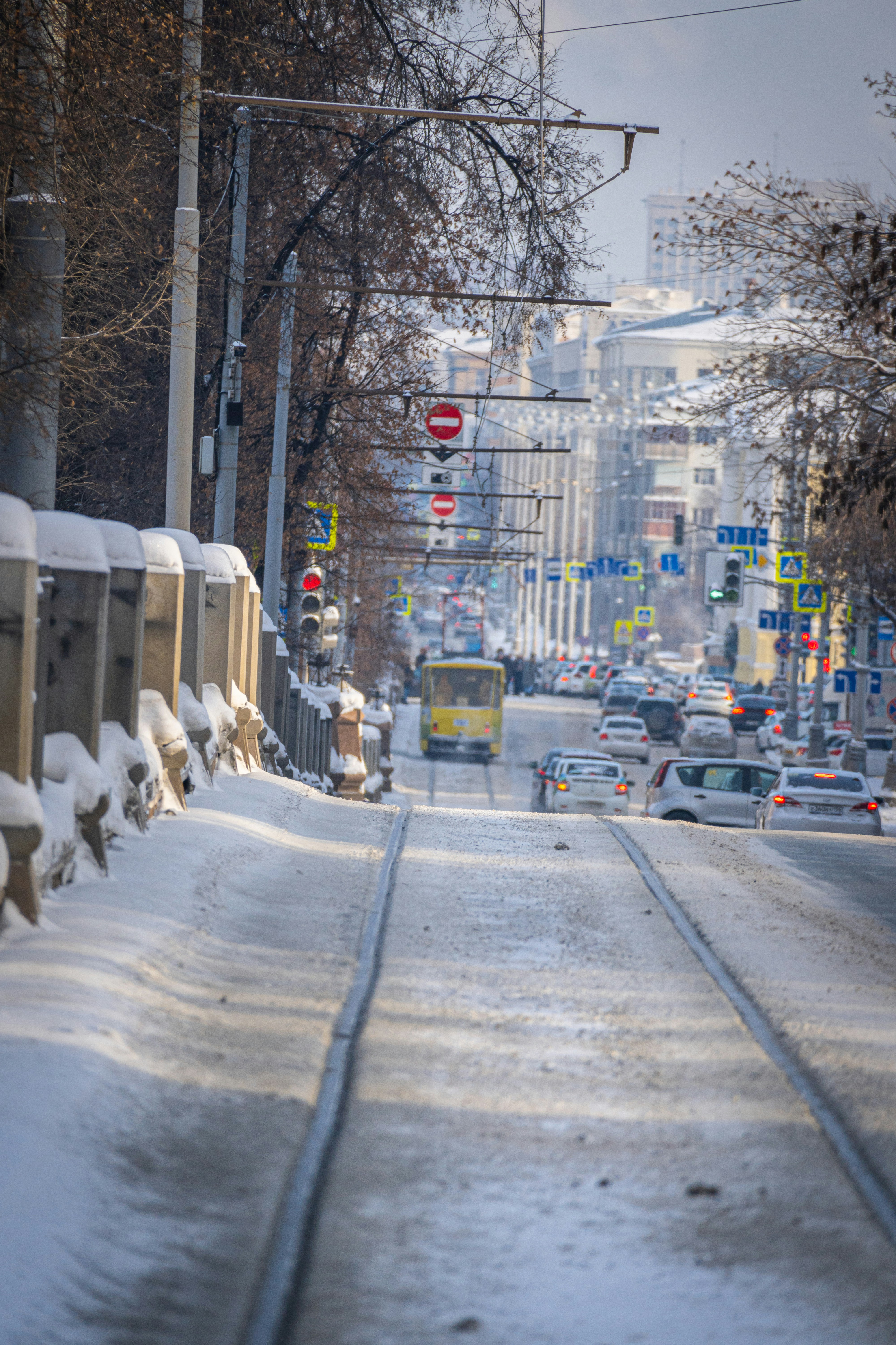 Snow-covered urban street with tram tracks, lined by traffic and streetlights under a cloudy sky.