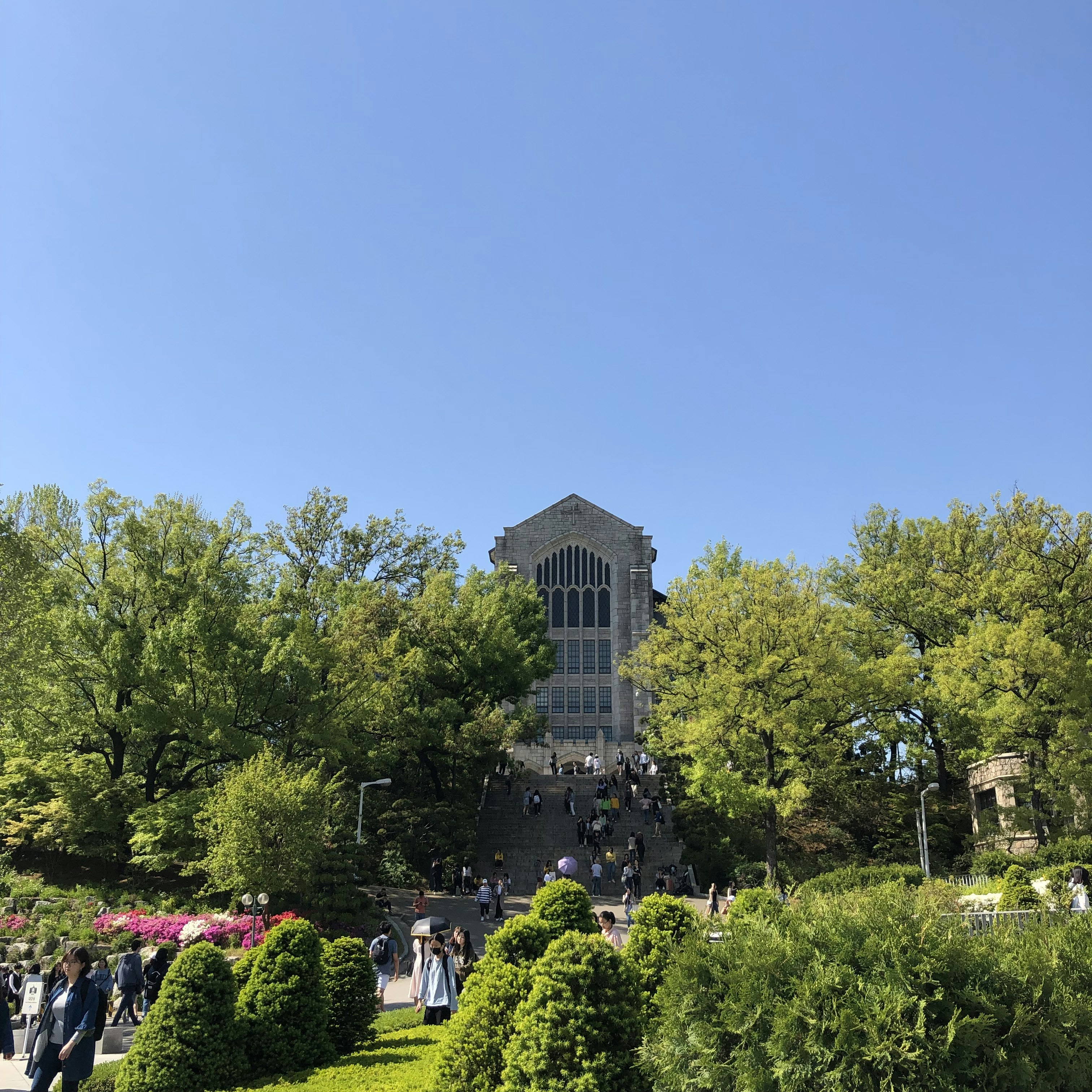 Visitors ascend a grand staircase framed by lush greenery and vibrant flowers, leading to a historic building under a clear blue sky.