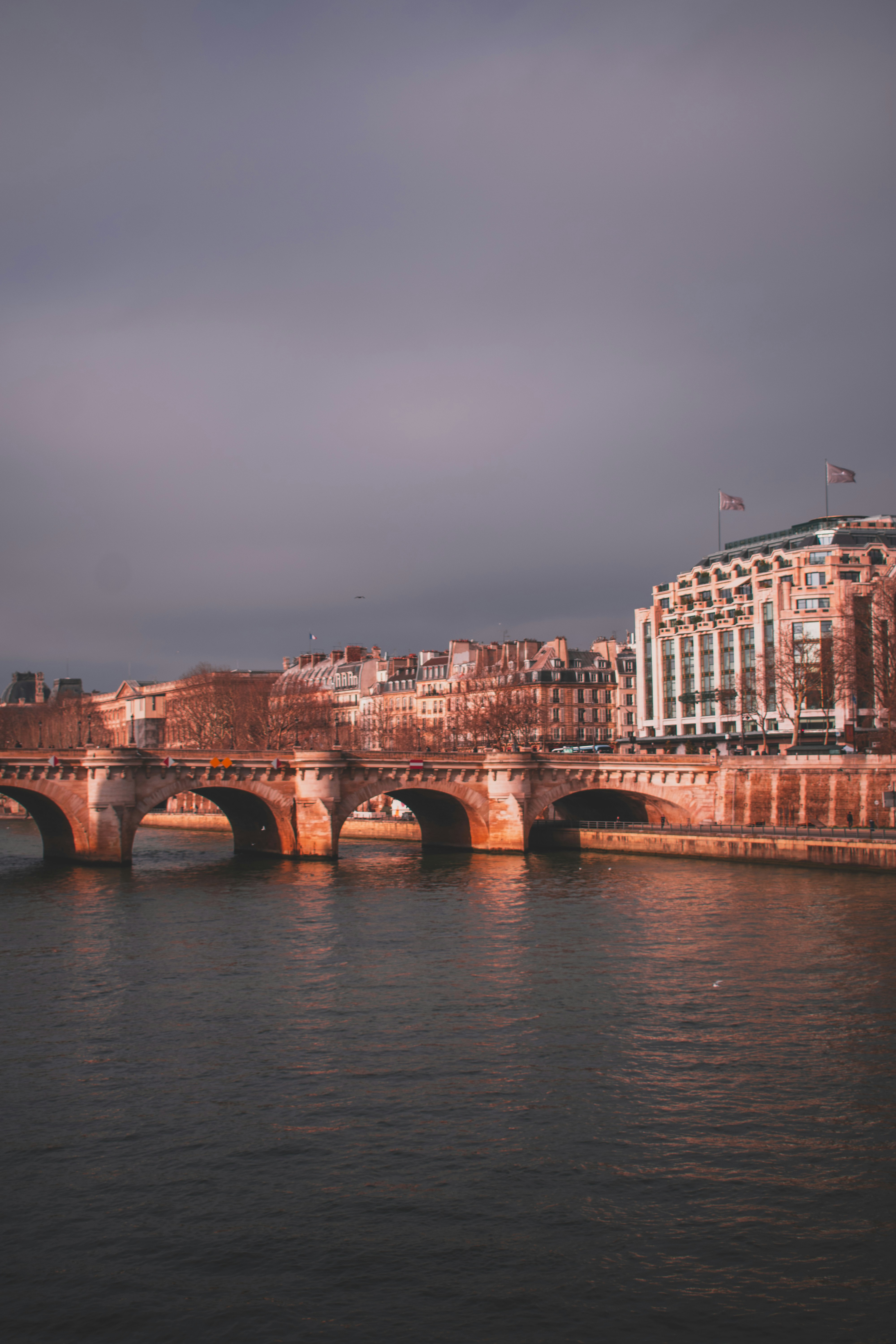 Historic stone bridge arches over tranquil waters, framed by modern architecture under a moody sky.