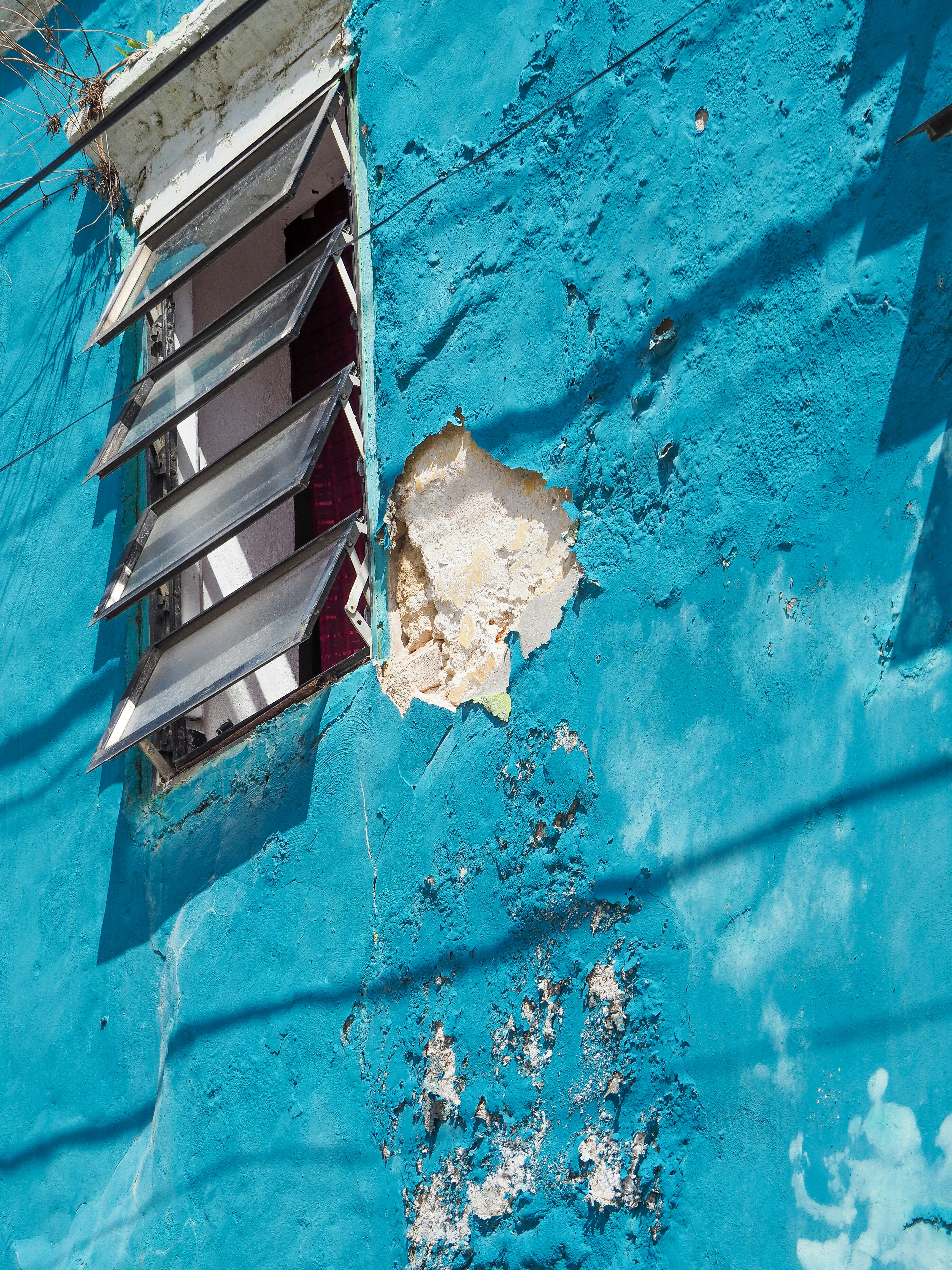 Weathered blue wall with peeling paint and a partially open window, revealing a glimpse of interior decor.