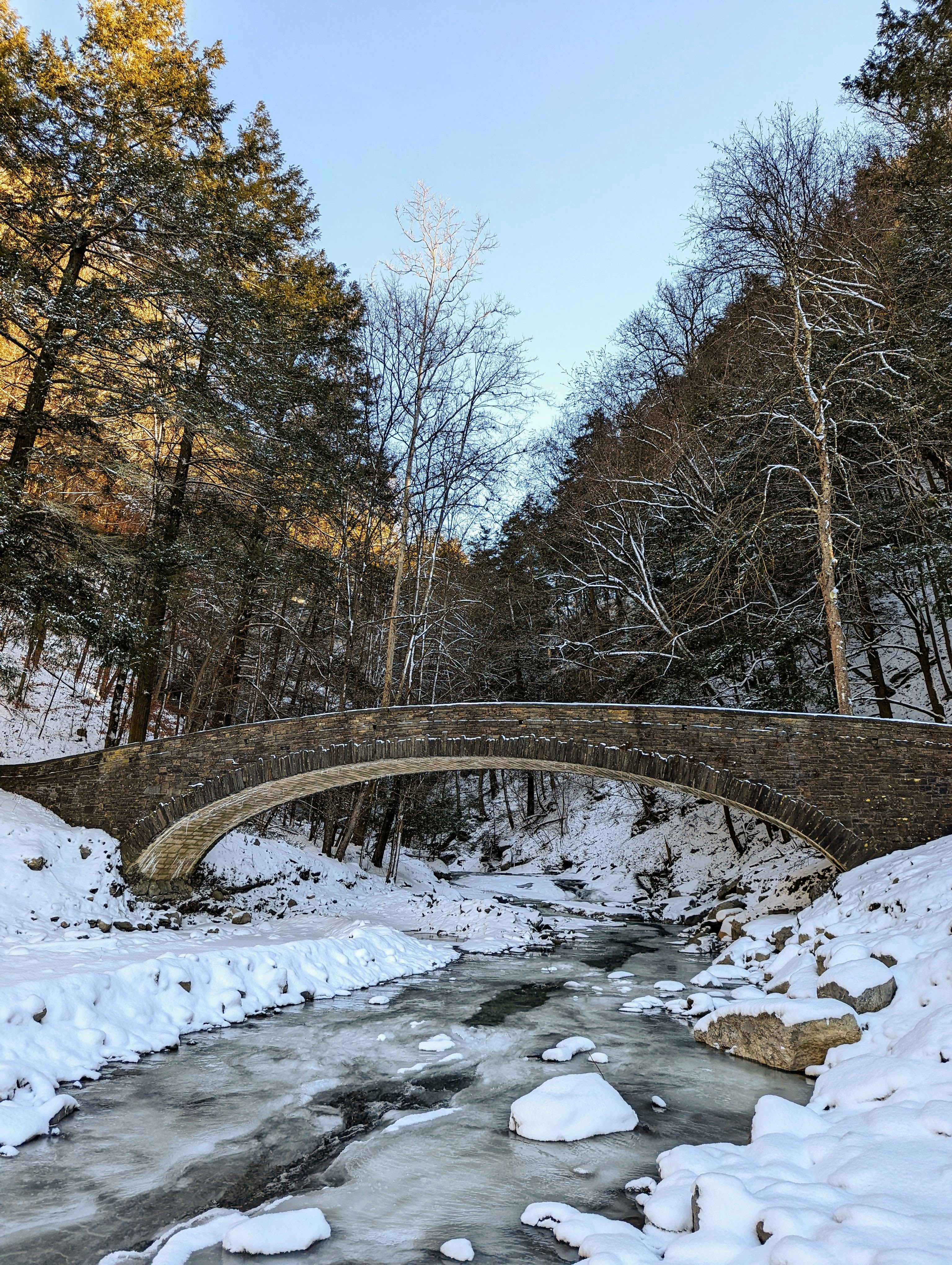 Photograph of a stone arch bridge spanning a partially frozen river, flanked by snow-covered pines under a clear blue sky.