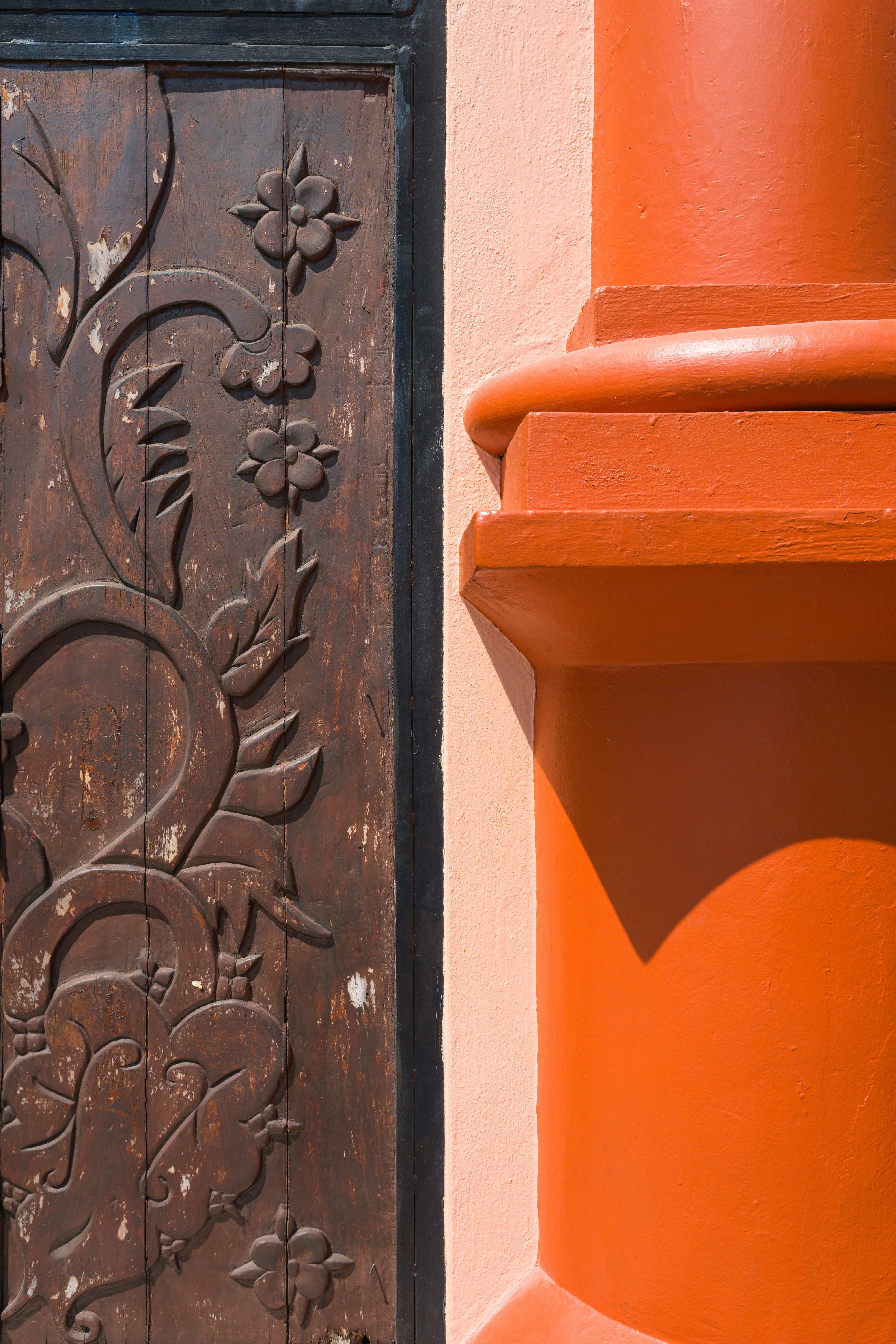 Close Up of Ornate Carving on Wooden Door and Orange Column - A close-up shot of a weathered wooden door with intricate floral carvings, positioned next to a bright orange column. The wood grain is visible on the door, and the floral carvings are detailed and elegant.