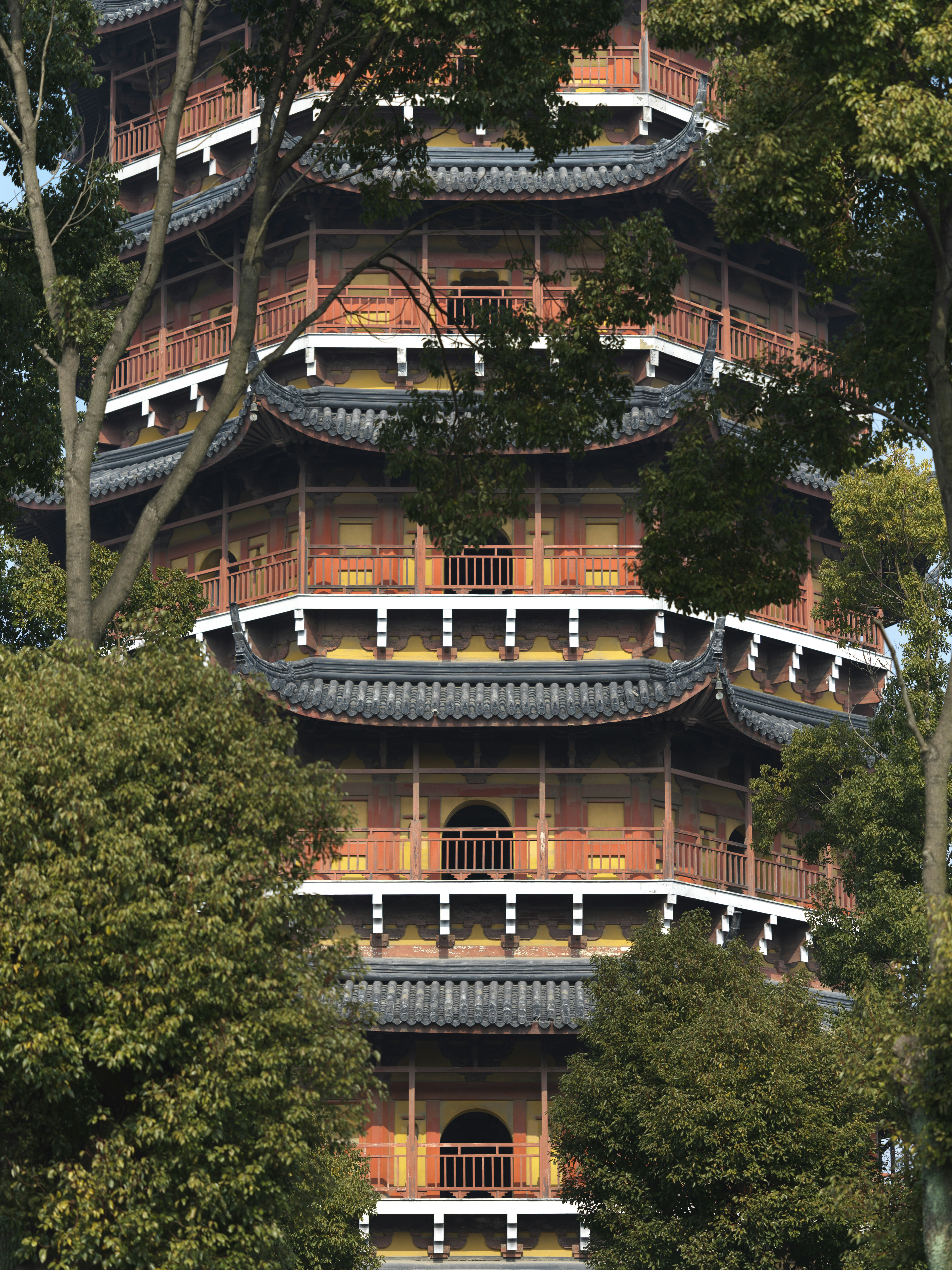 Multi-tier Chinese pagoda rises through green trees in a tranquil park, its red walls and dark tiled roofs repeating along each balcony. Photograph capturing architectural rhythm and natural framing.
