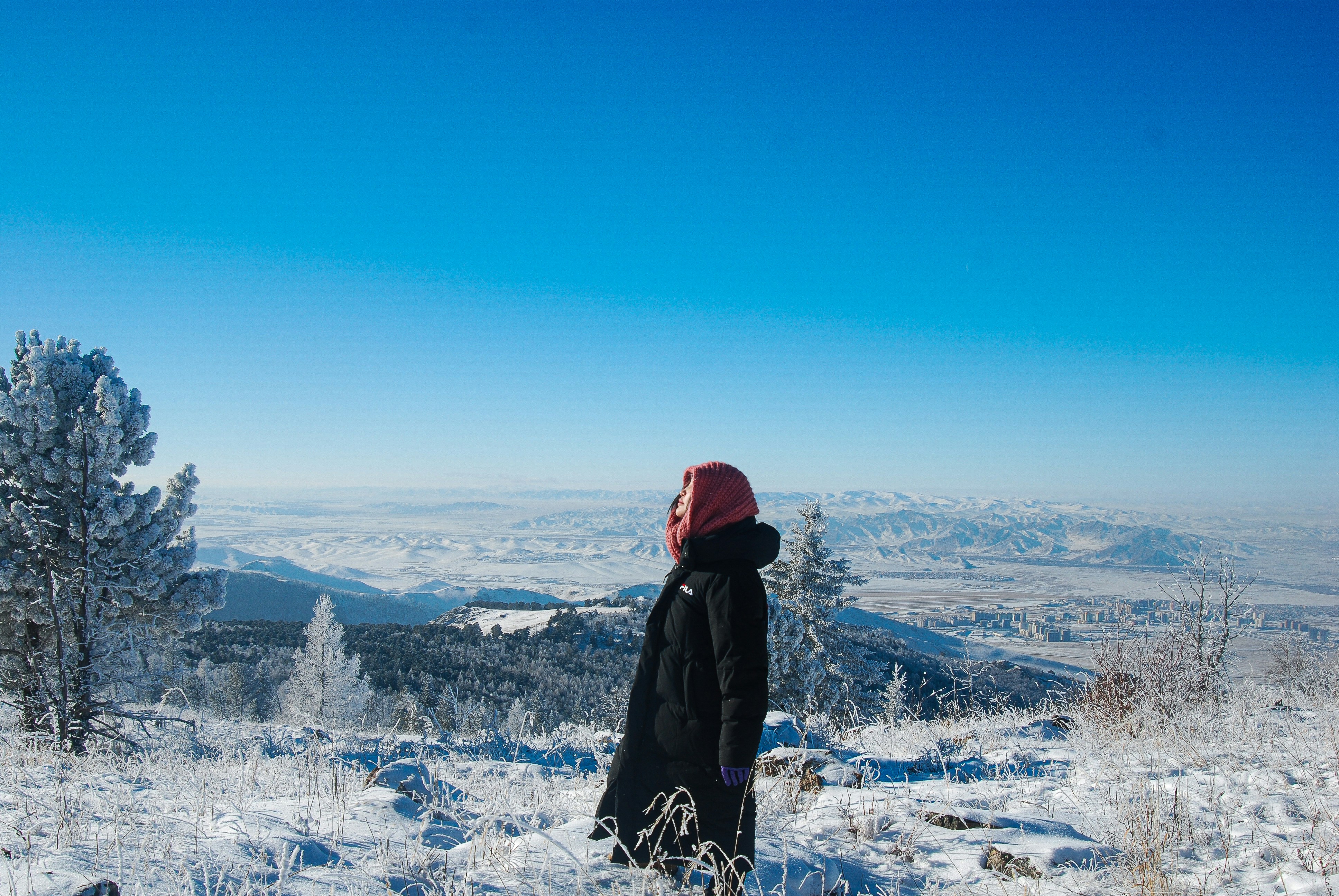 Person in winter clothing stands amidst snowy landscape with expansive mountain views under a clear blue sky.