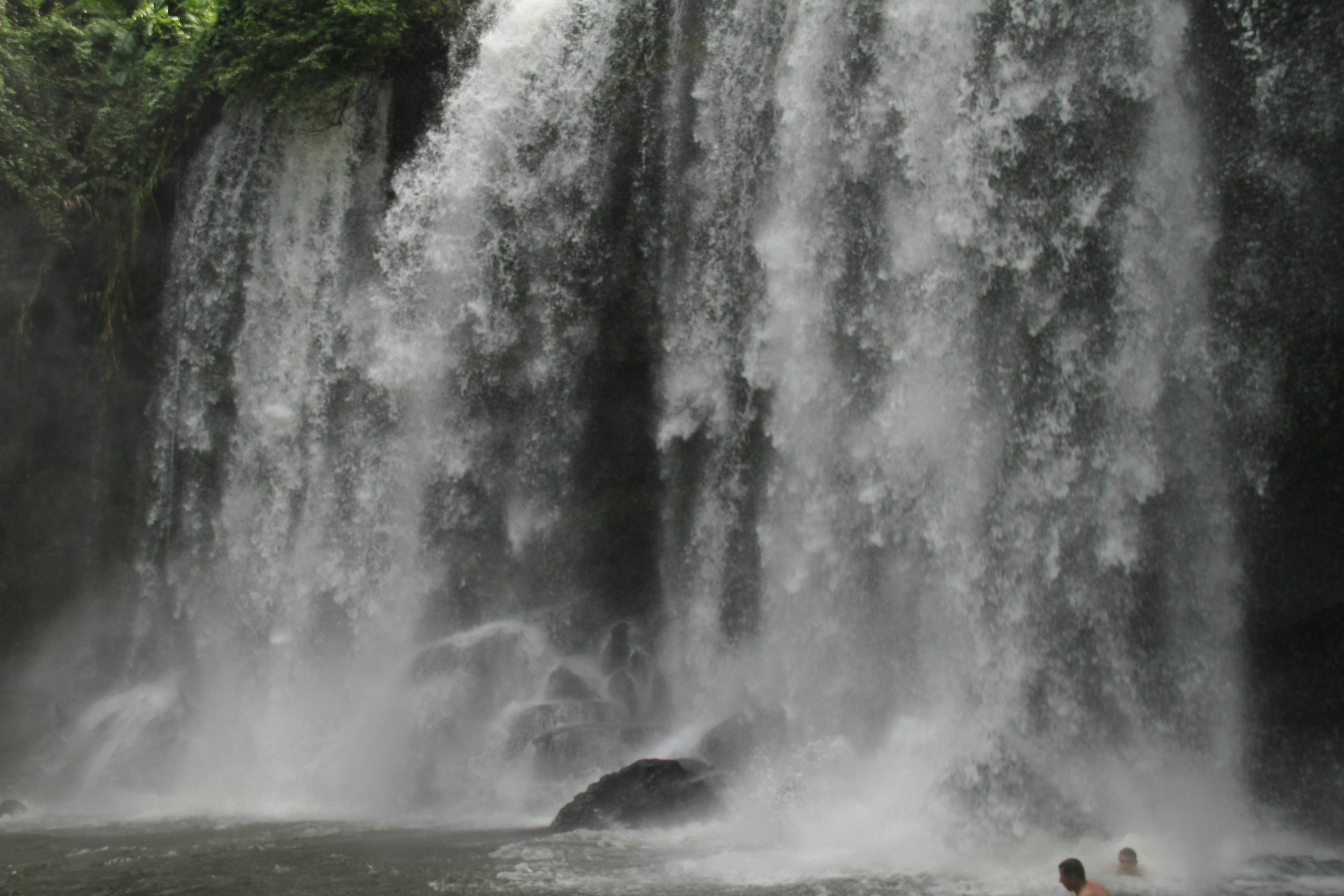 Powerful waterfall cascading down rocky cliffs, with two figures enjoying the mist in the foreground. The lush greenery frames the scene beautifully.