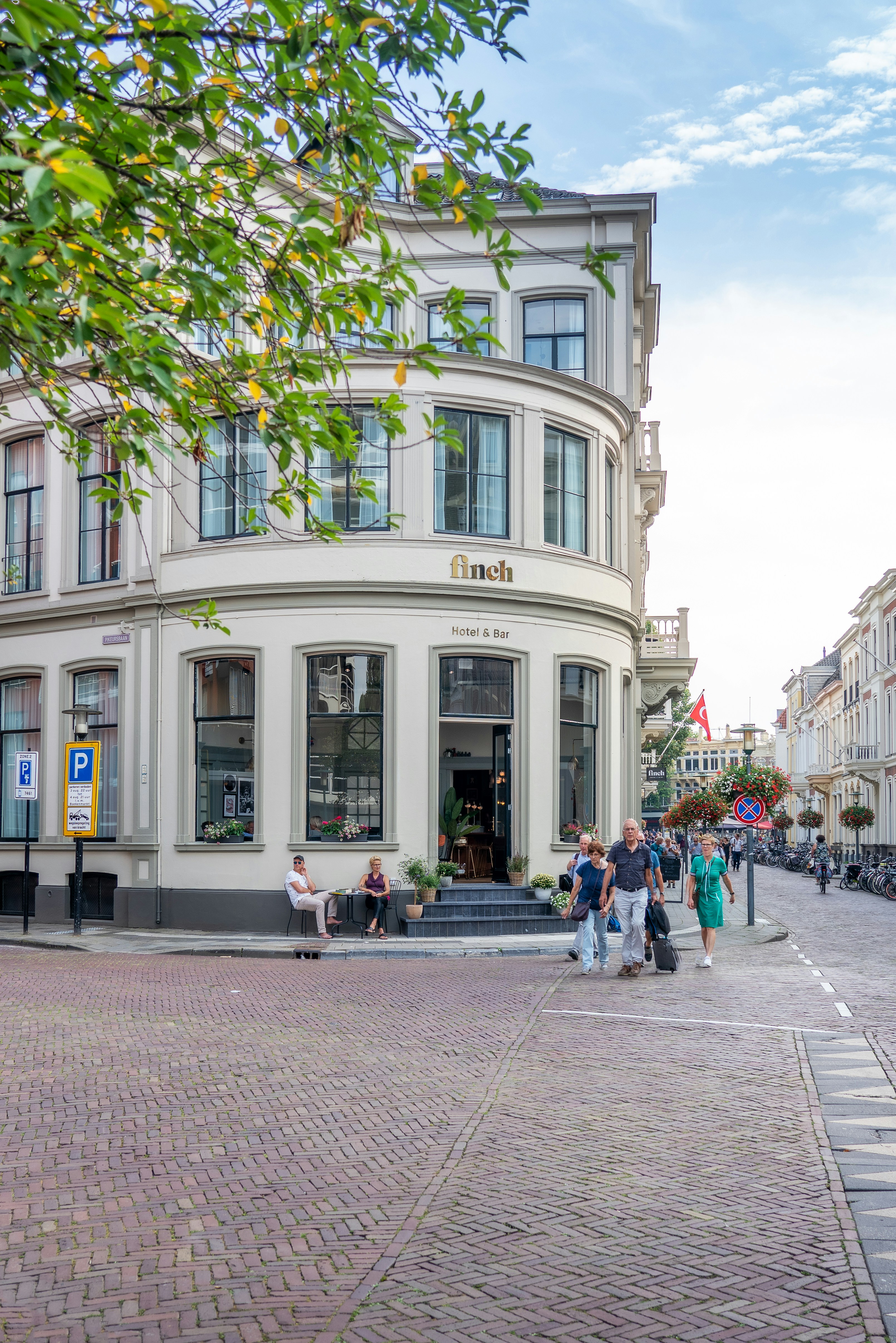 Charming hotel facade featuring a welcoming entrance, surrounded by pedestrians and lush greenery. The scene captures the essence of urban life.