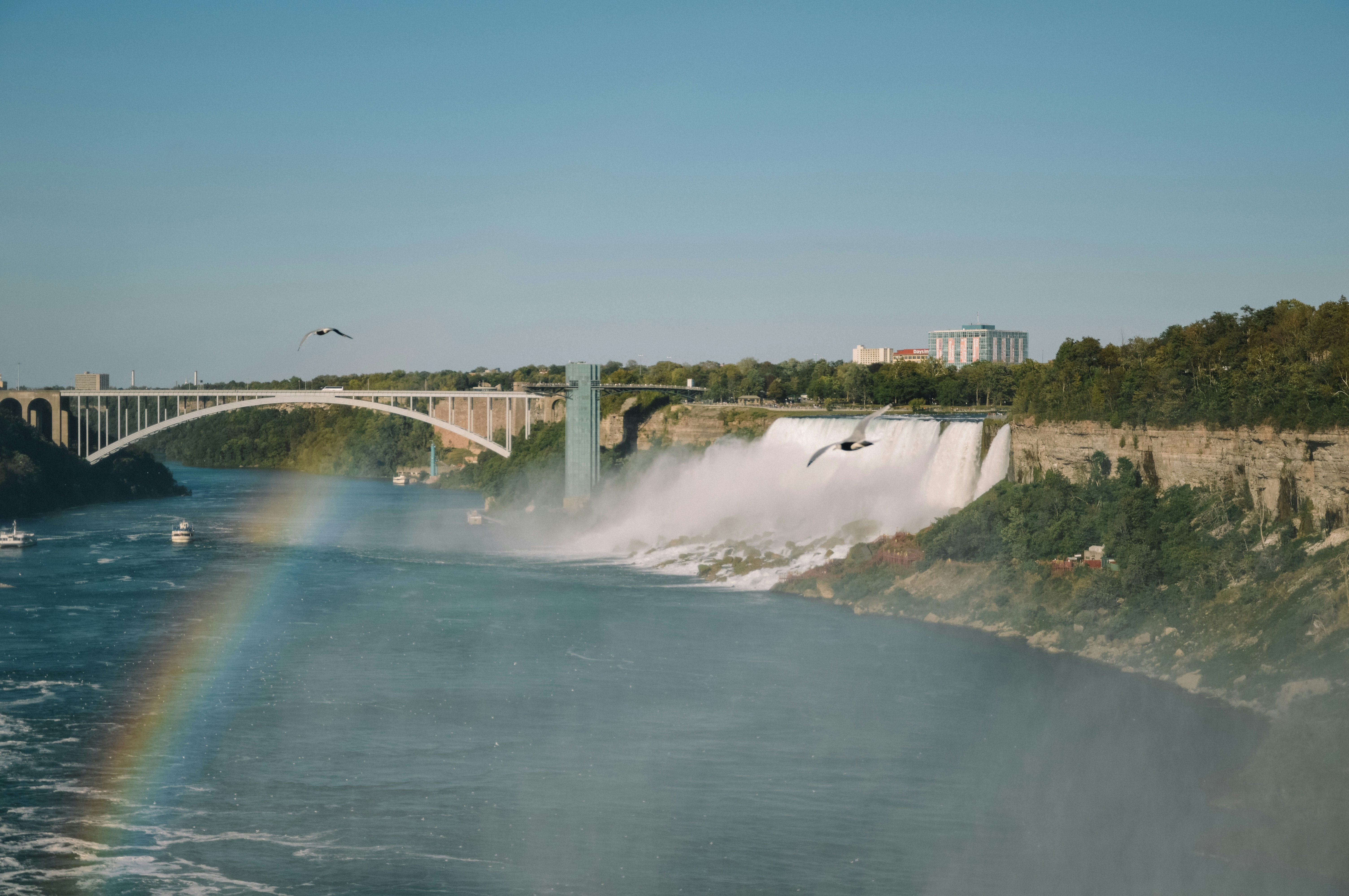 A vibrant rainbow arcs over the rushing waters of a majestic waterfall, framed by a bridge and lush greenery. The scene captures the essence of natural beauty and tranquility.