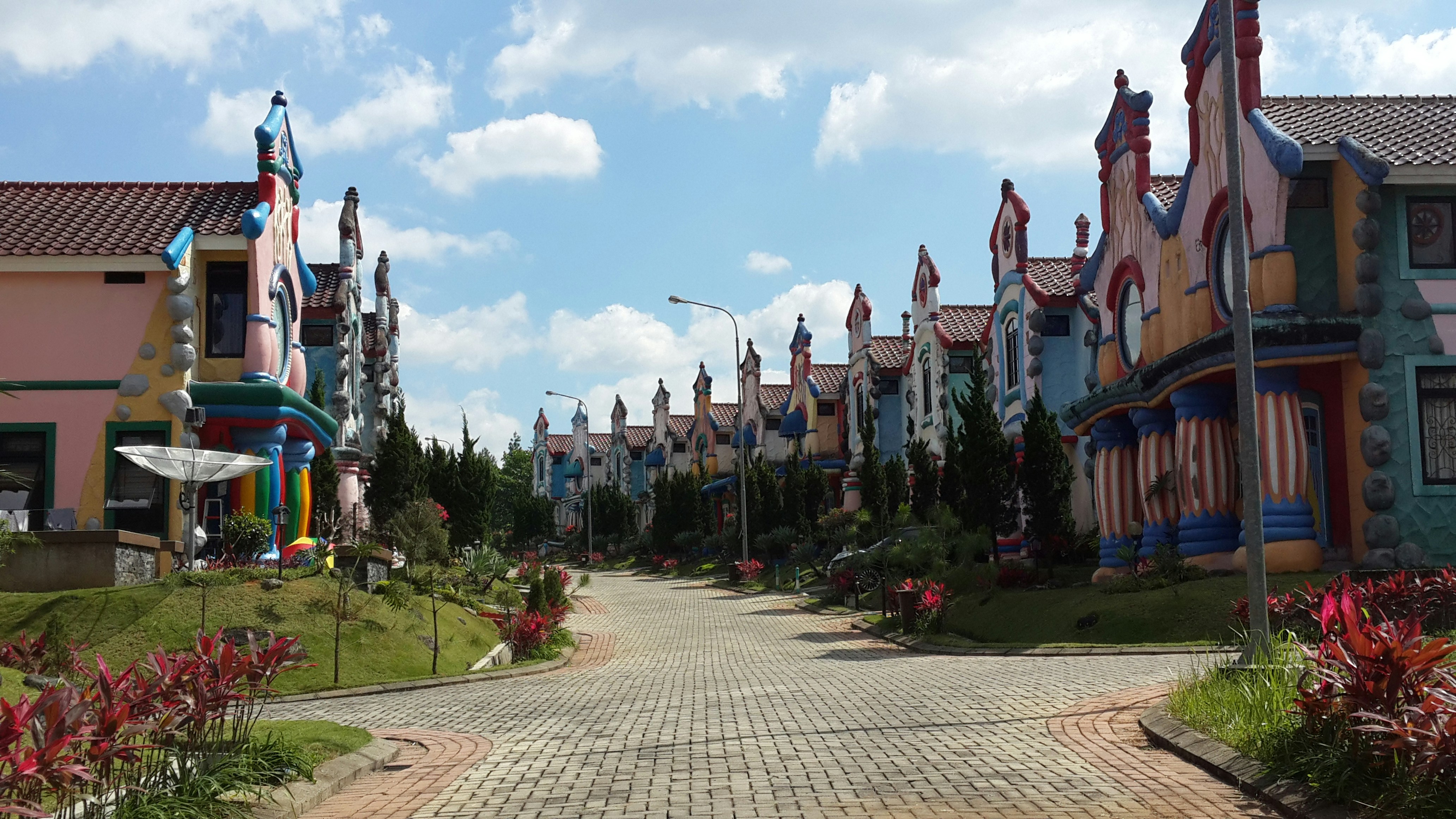 A vibrant street lined with uniquely designed houses featuring playful architectural elements and colorful facades. Lush greenery and flowers enhance the charming atmosphere.