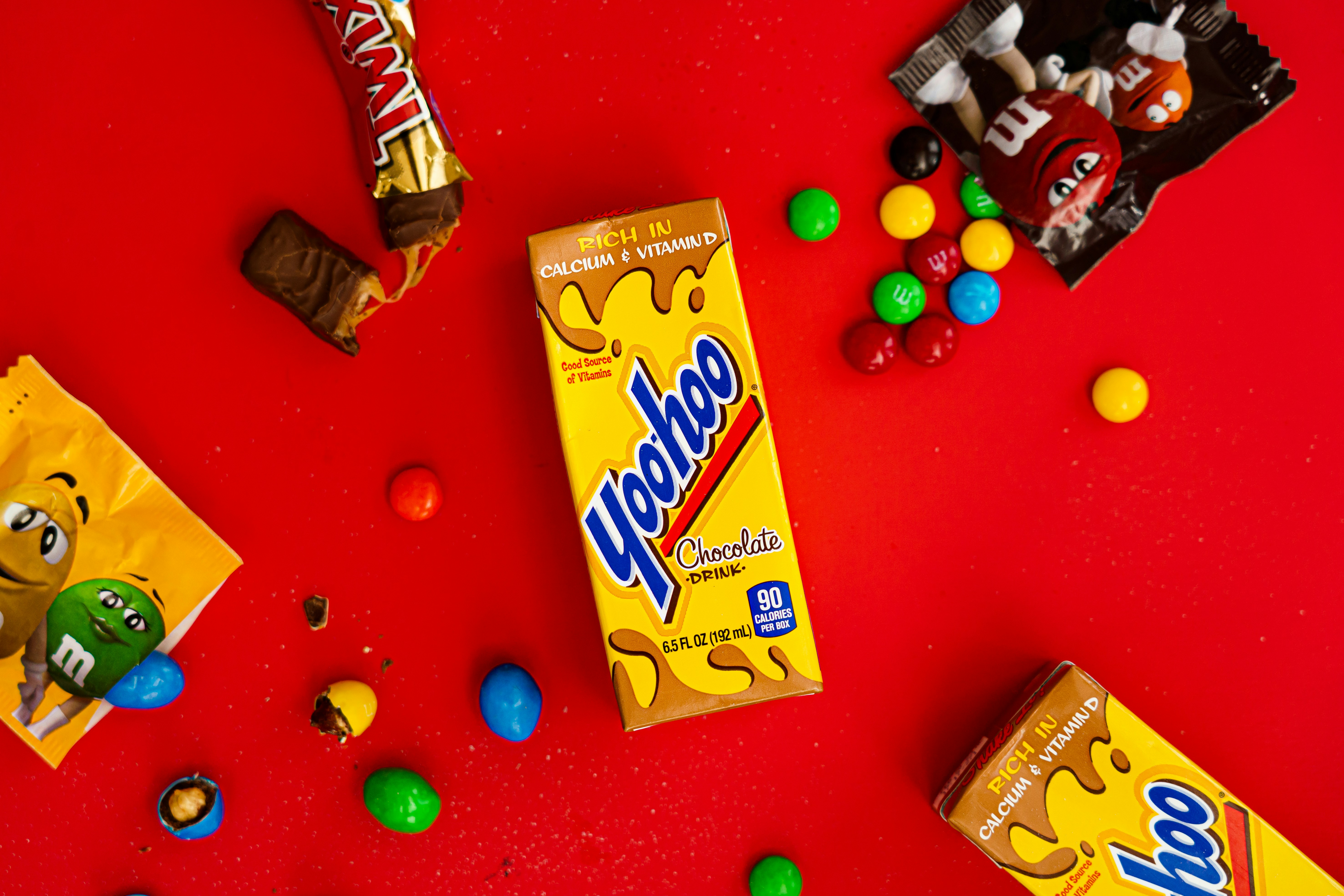 Colorful candies and chocolate bars scattered around a Yoo-hoo chocolate drink box on a vibrant red background.